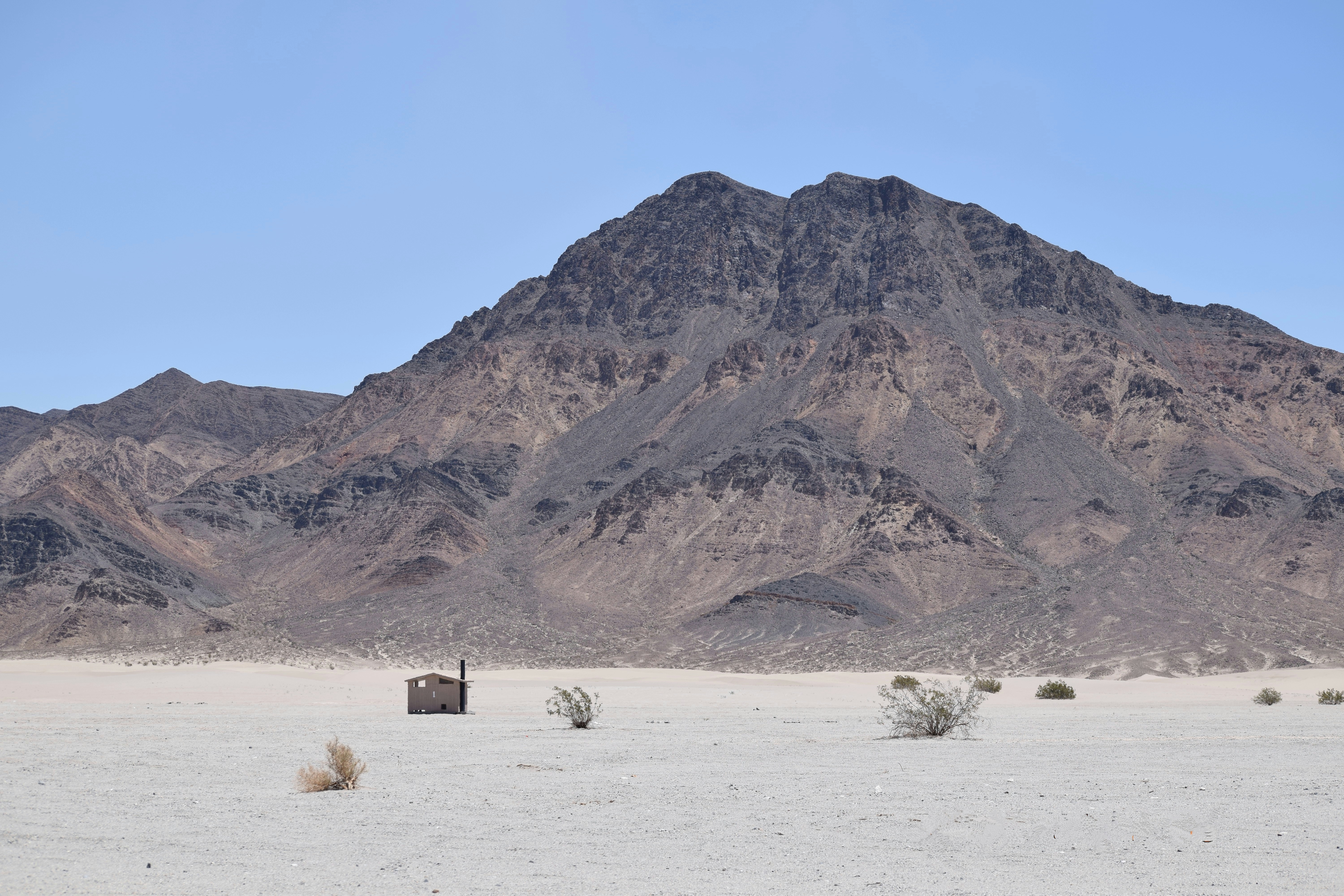 A solitary building stands in the vast desert expanse of Death Valley with rugged mountains rising in the background under a clear blue sky.