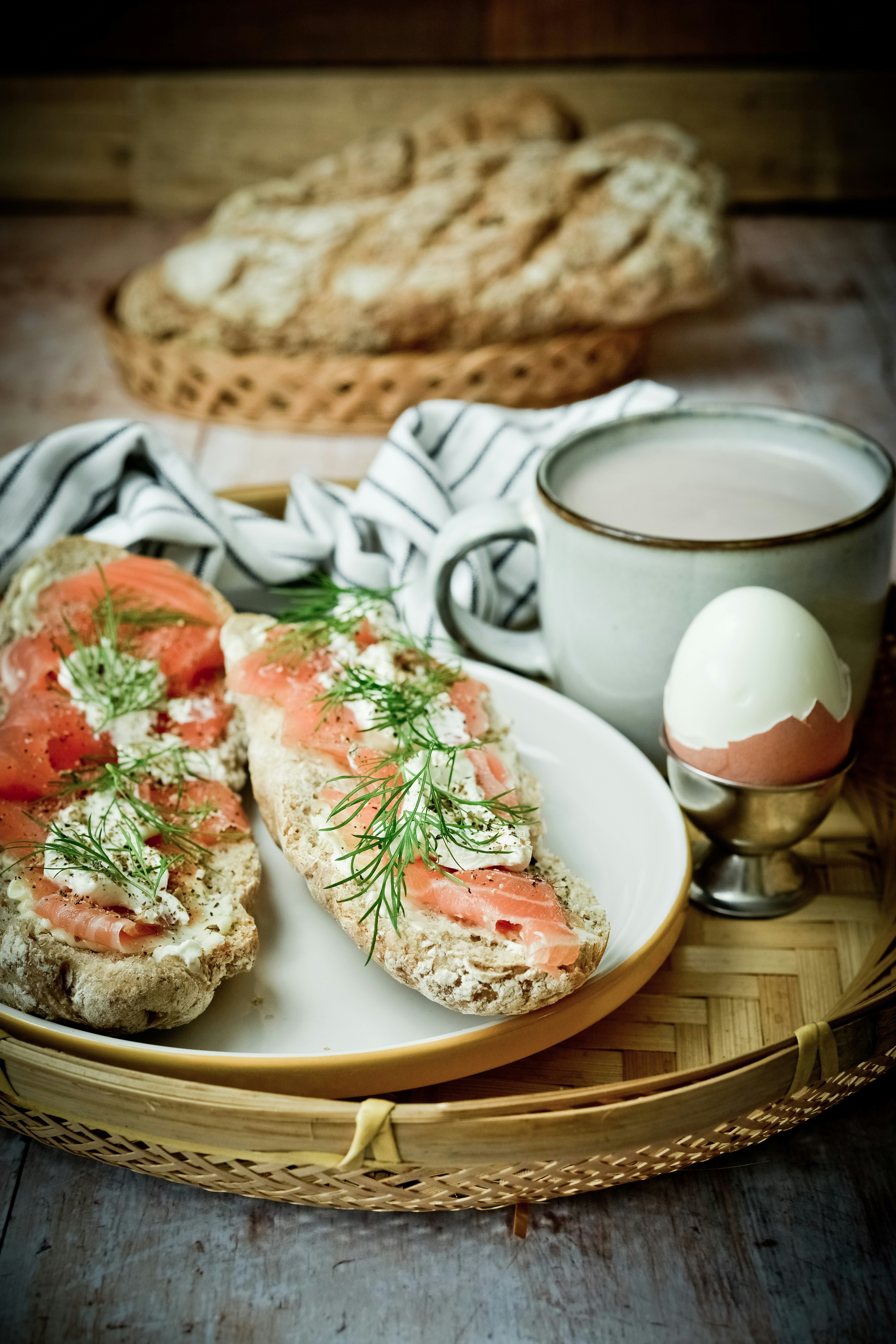Artfully arranged open-faced sandwiches topped with smoked salmon and fresh dill, accompanied by a soft-boiled egg and a warm beverage in a rustic setting.