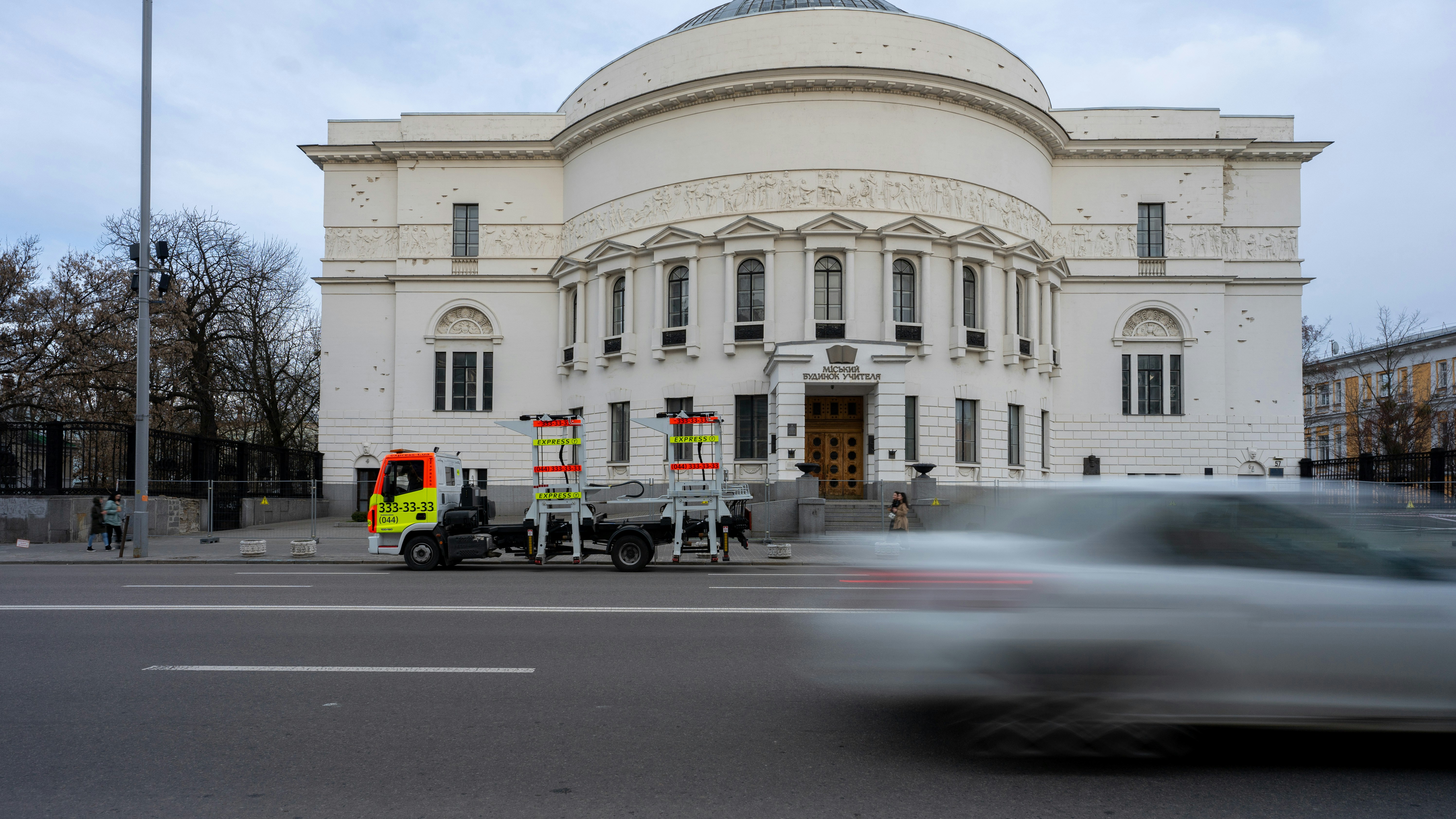 Tow truck parked near a neoclassical building as a car blurs by on the street.