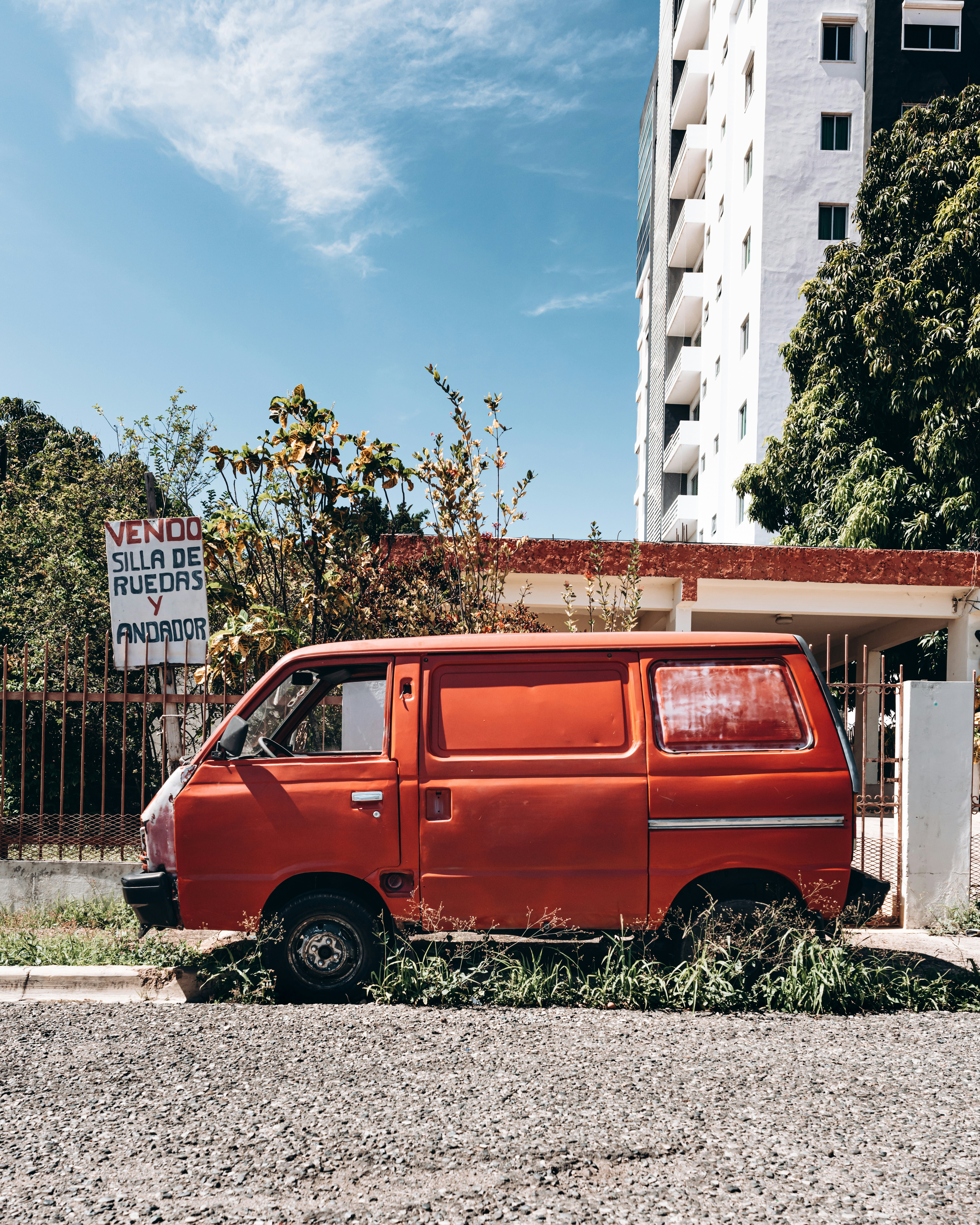 Red mini van parked beside a tall building and overgrown grass under a bright blue sky.