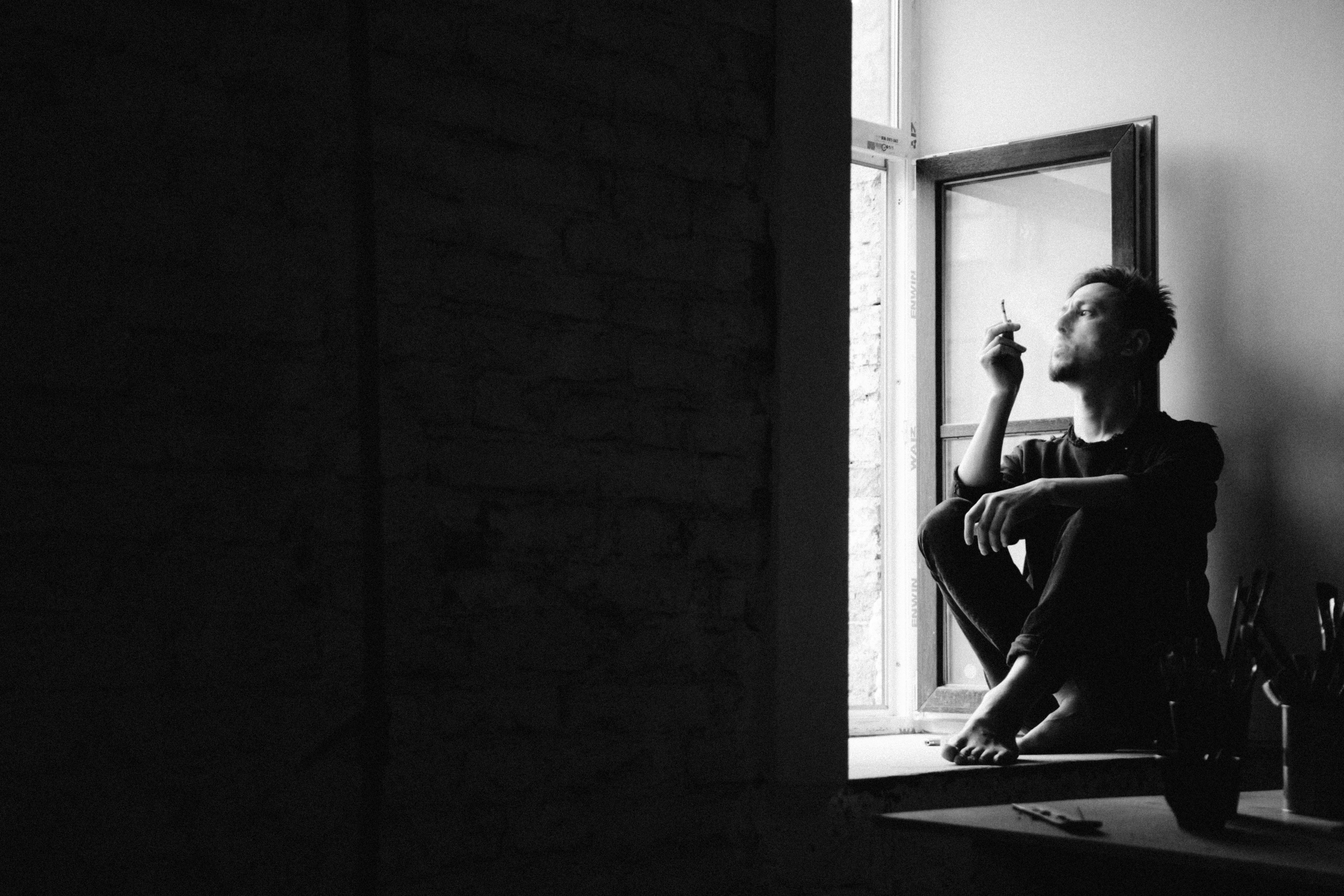 A contemplative figure seated in a window nook, exhaling smoke while surrounded by soft light and shadows.