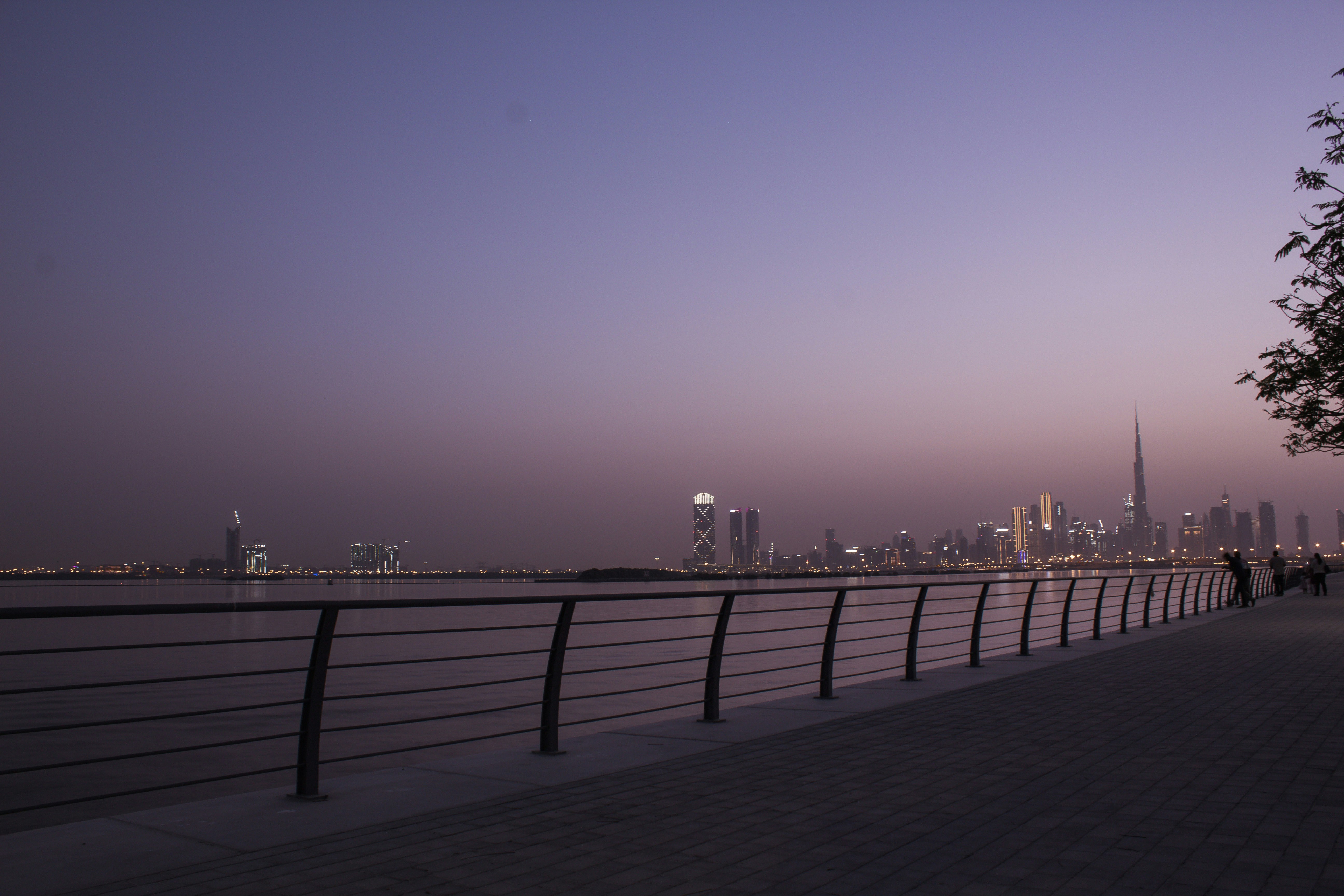 City skyline illuminated at dusk, reflecting on calm waters with a modern promenade in the foreground.