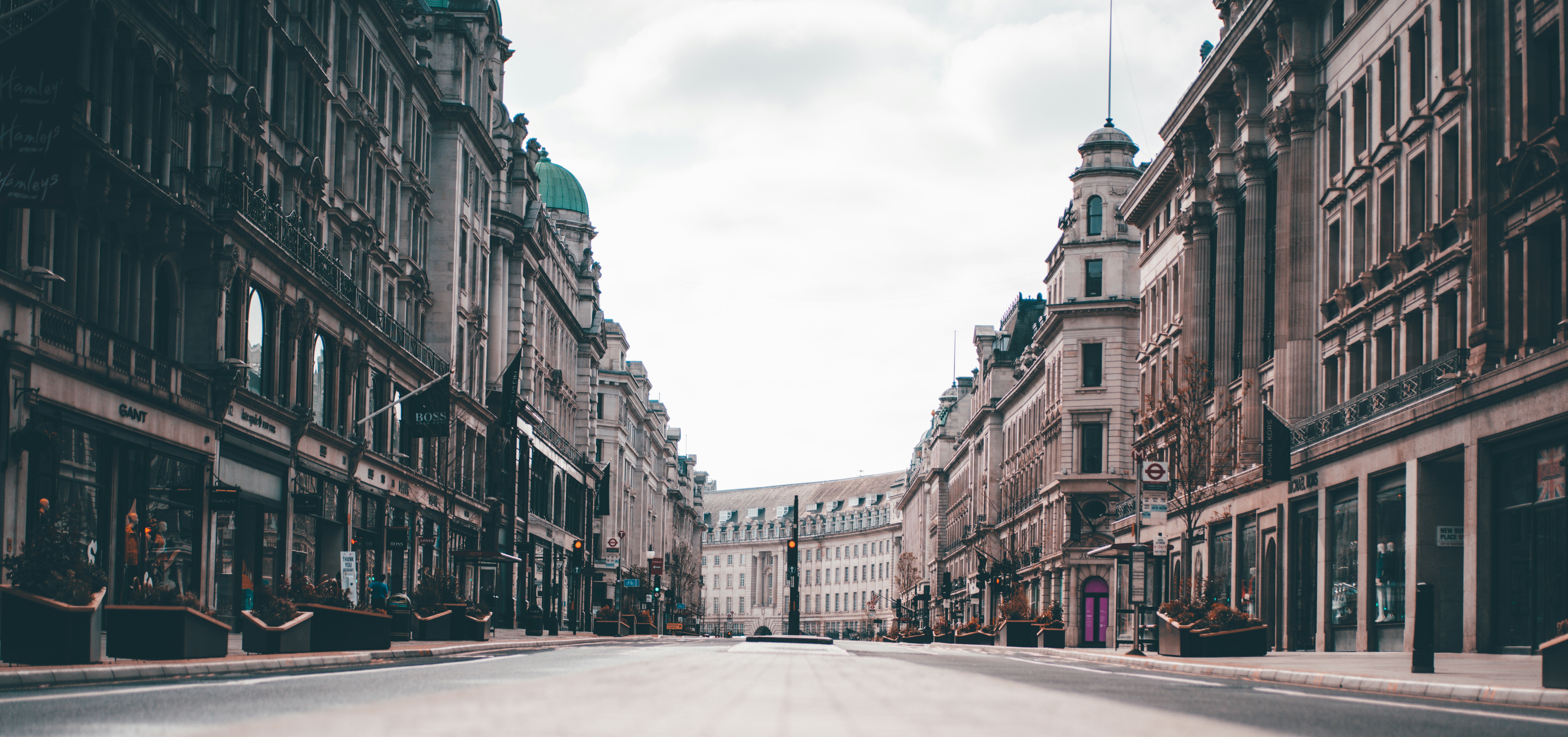 This captivating image showcases a deserted city street flanked by grand, historic buildings under a moody, overcast sky. The composition draws the eye towards the vanishing point, emphasizing the symmetry and scale of the architecture. Muted tones of grey and beige dominate the scene, while soft lighting enhances the peaceful atmosphere, making it a visually striking representation of urban tranquility.