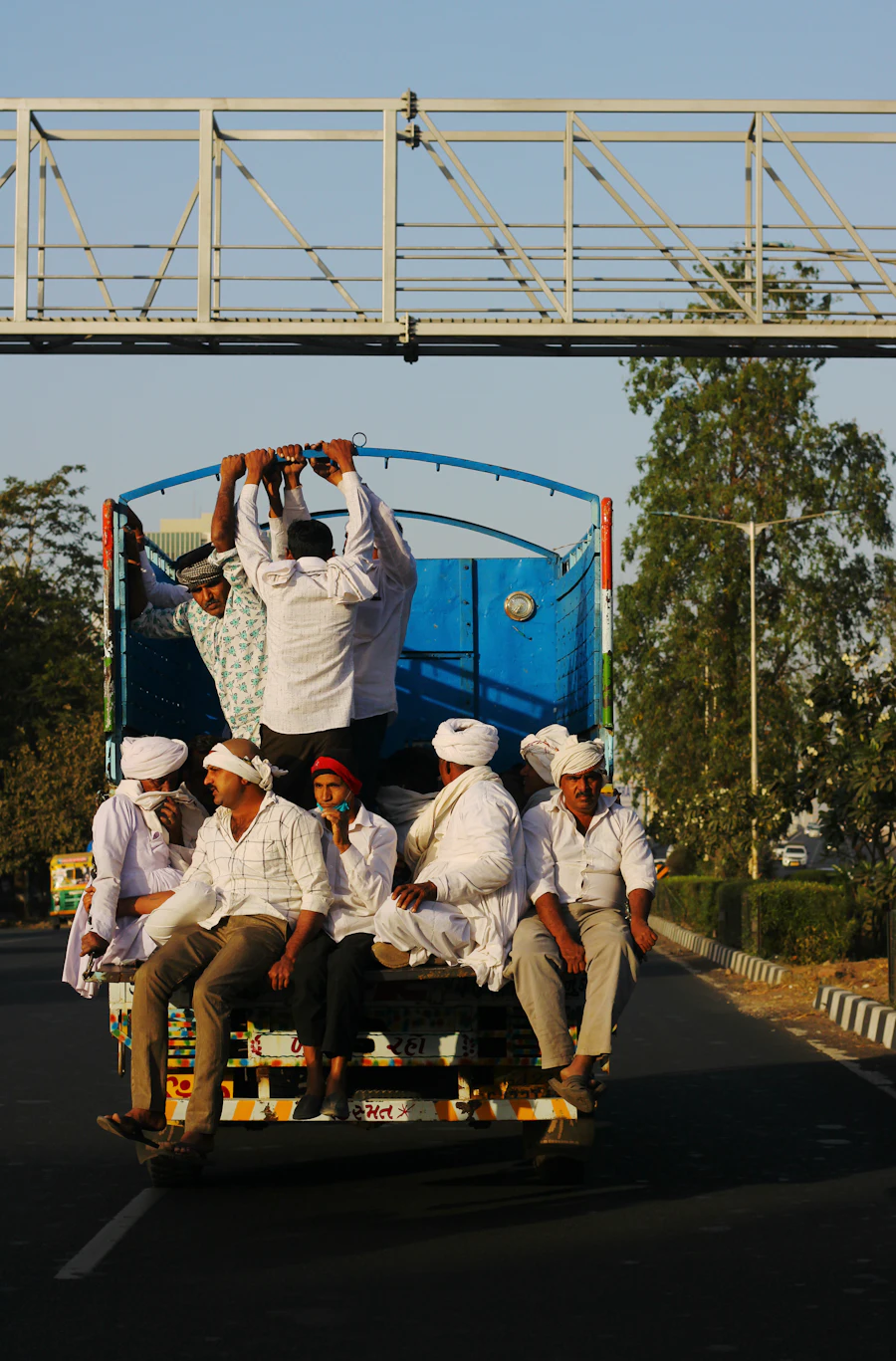 A group of men in white clothing and turbans ride on the back of a blue truck on a sunlit road, with an overhead bridge framing the scene. Trees and roadside greenery line the background.