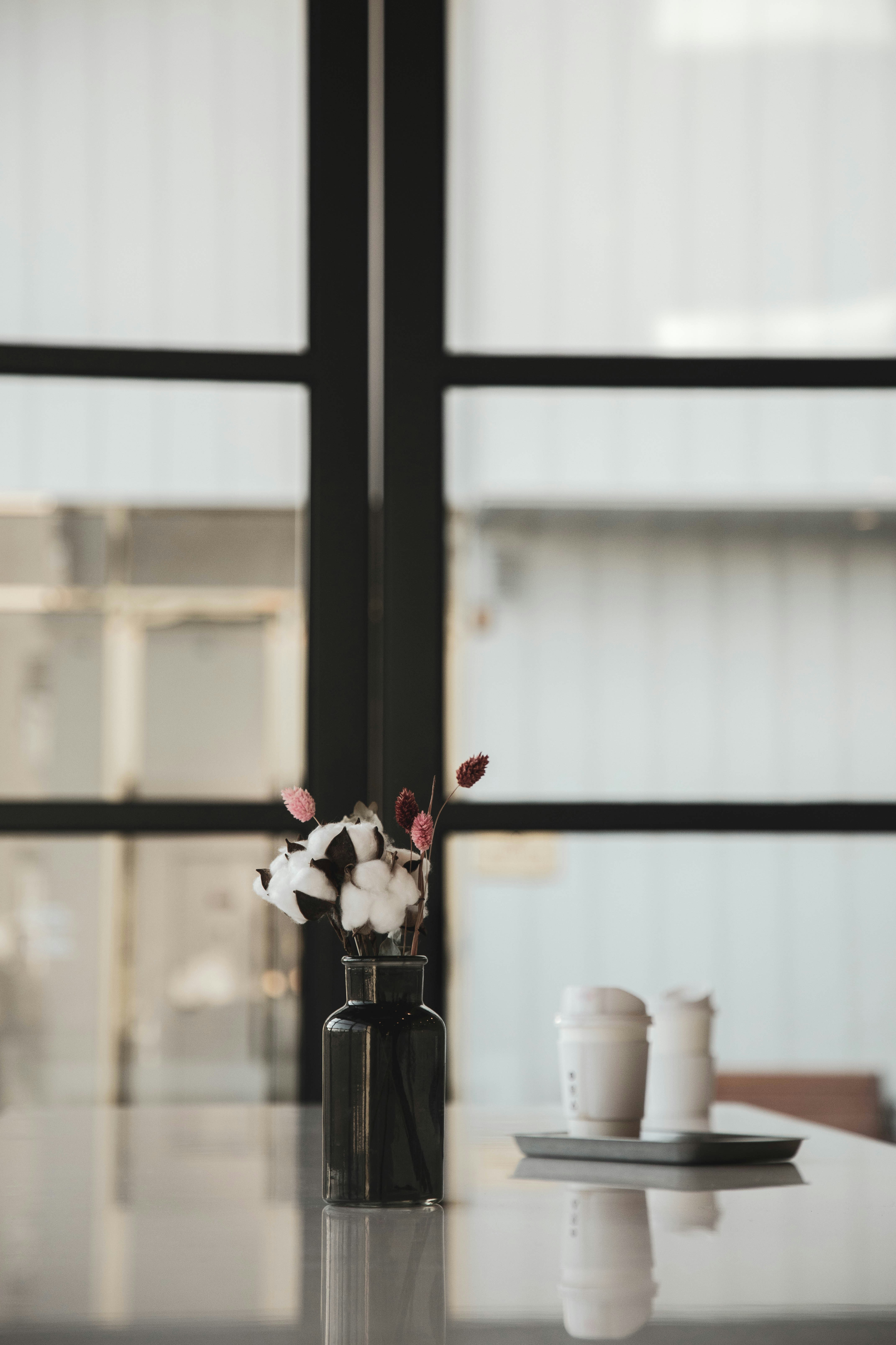 Minimalist arrangement of cotton flowers in a black vase on a table, with coffee cups in the background. Soft natural light enhances the serene atmosphere.