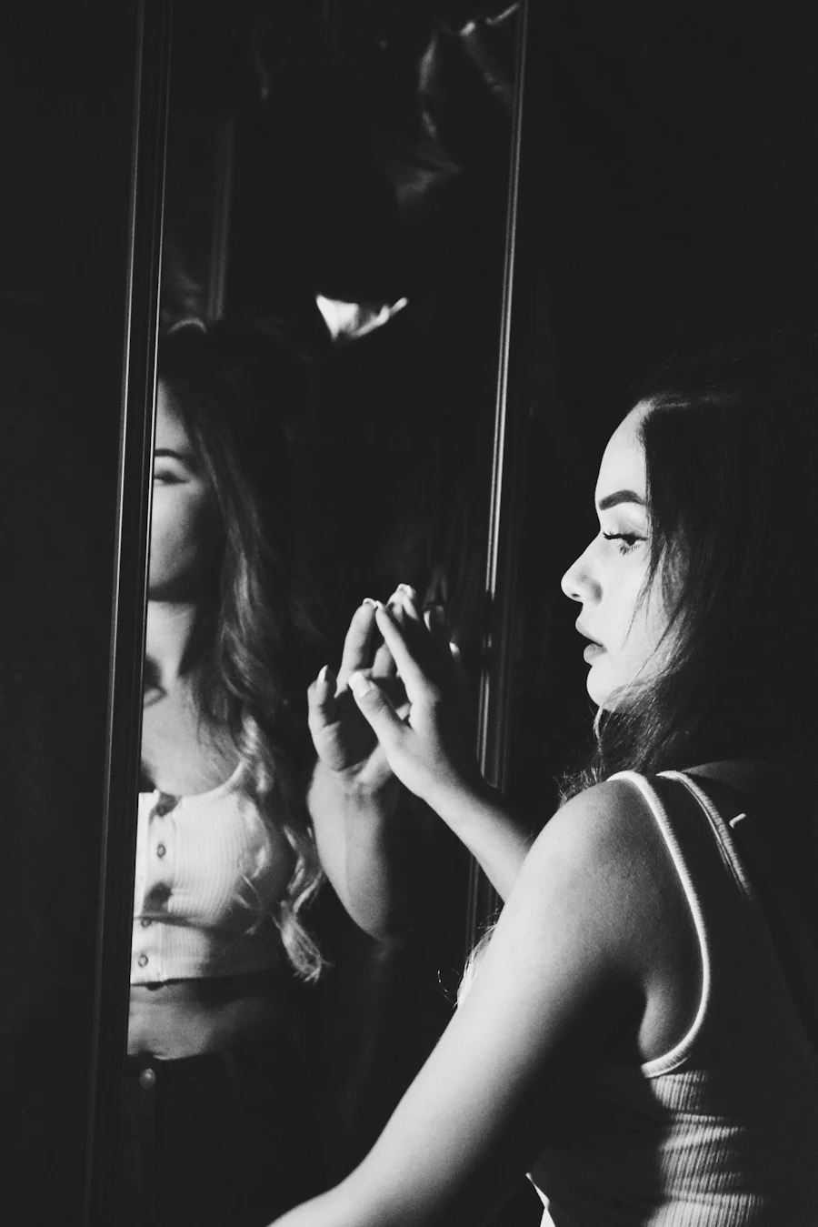Black-and-white portrait of a woman in profile beside a mirror, with her reflection visible on the left. Soft light highlights her face, hand, and shoulder against a dark background.