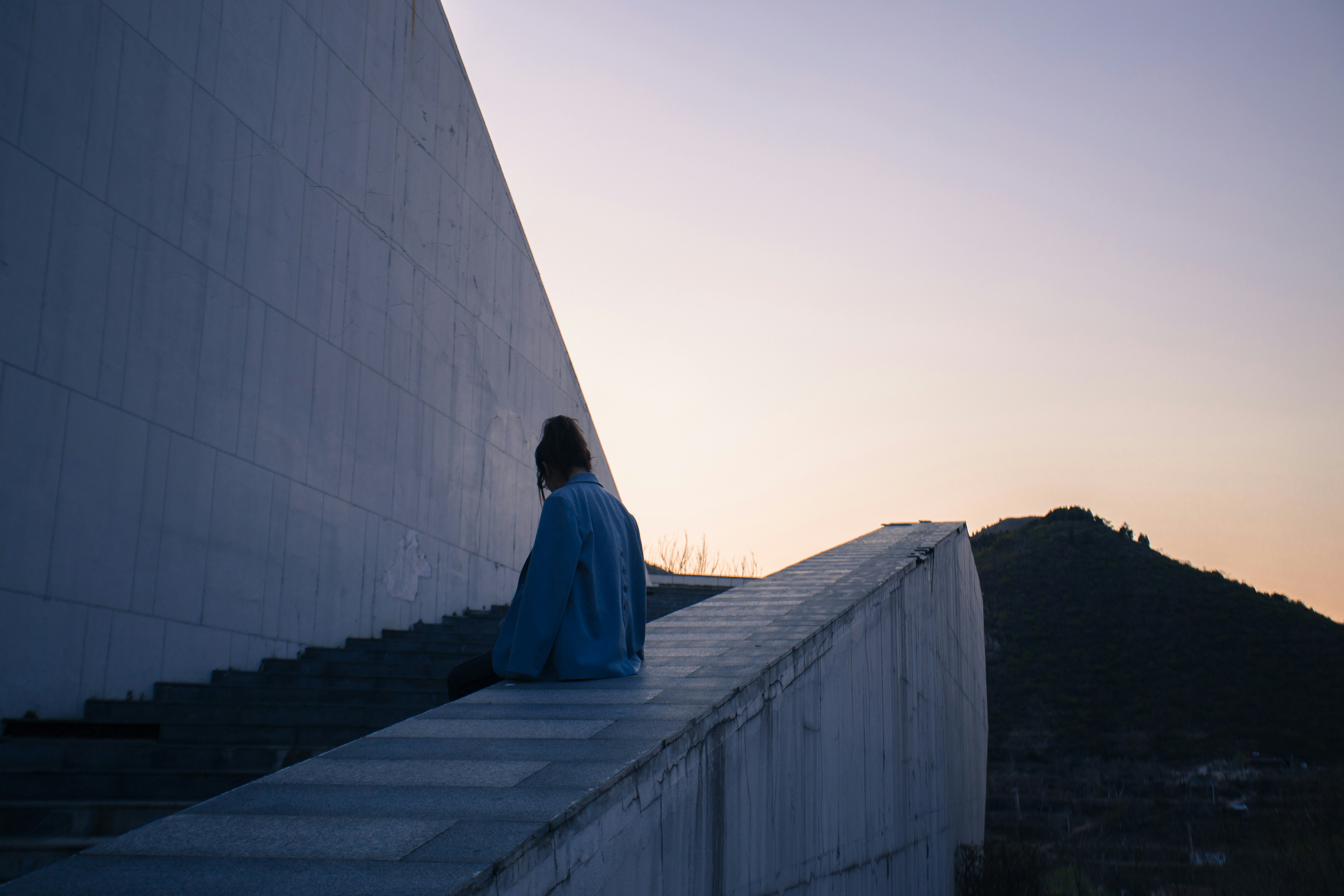 A figure in a blue coat sits contemplatively on a modern staircase, framed by a minimalist structure and a distant hill at dusk.