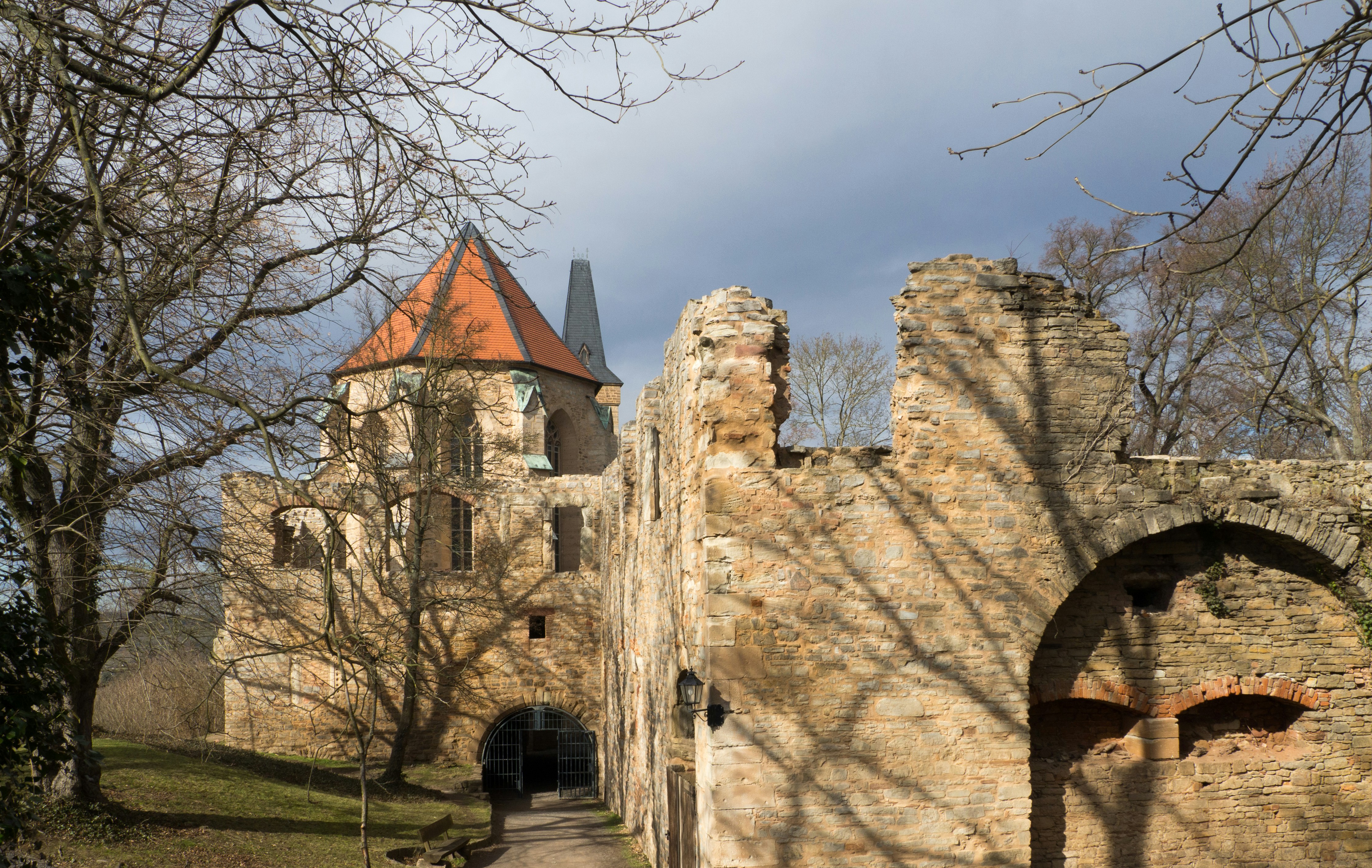 Ancient stone ruins with a striking red-roofed tower surrounded by bare trees and shadows. The scene evokes a sense of timelessness and history.