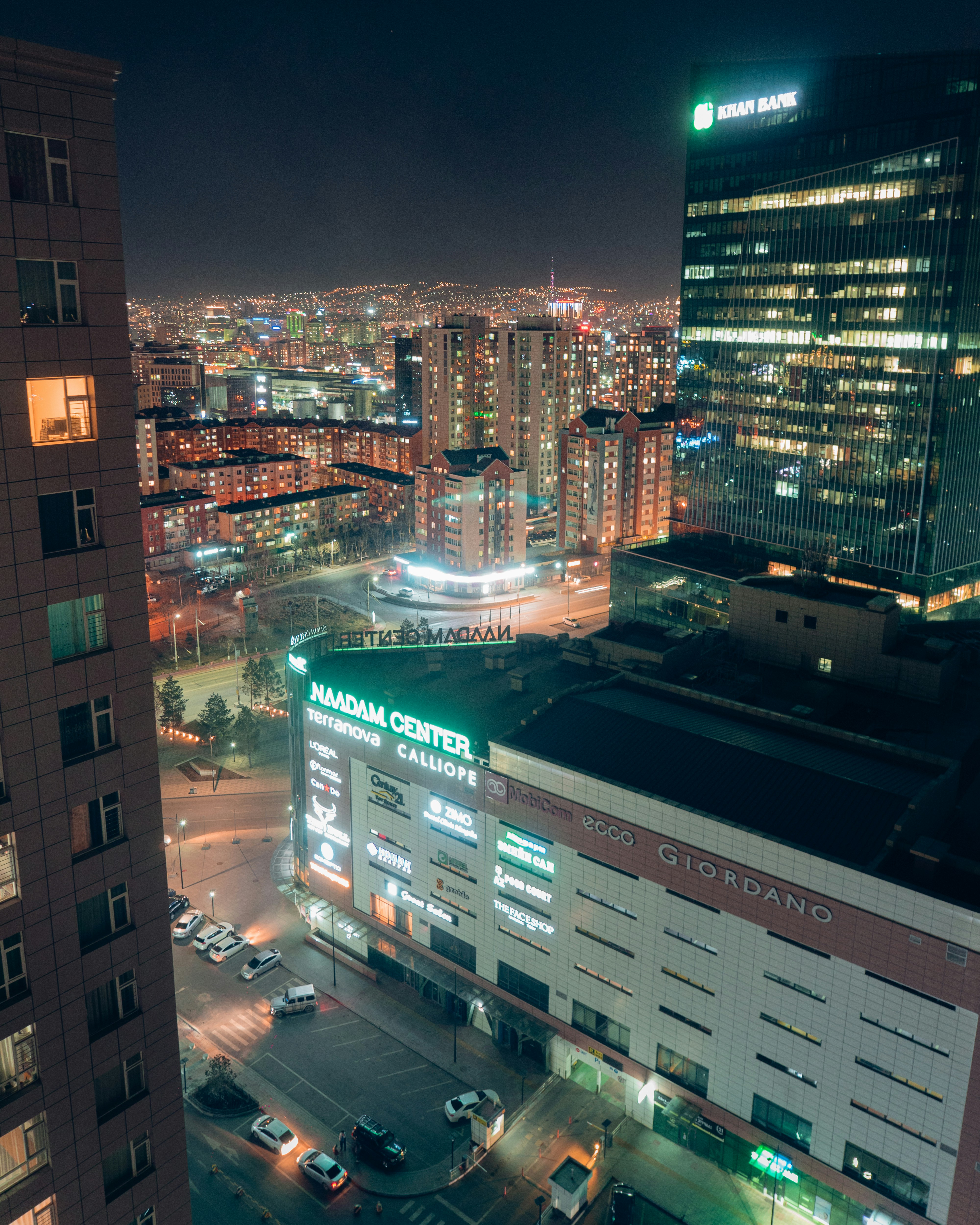 Vibrant city skyline illuminated at night, showcasing a blend of modern architecture and bustling streets. Neon signs highlight commercial spaces below.