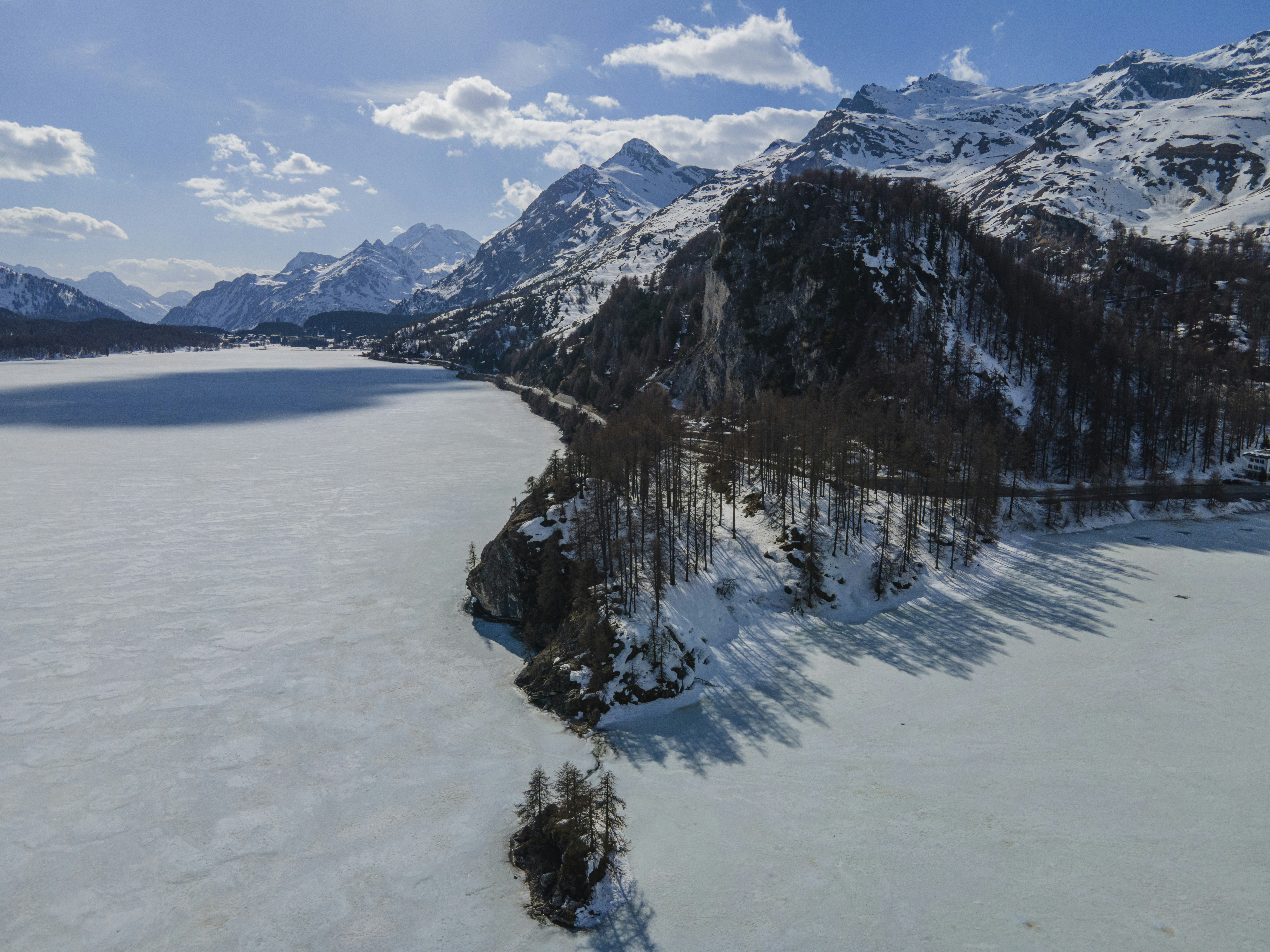 Snow-covered landscape with a frozen lake bordered by mountains and trees, showcasing the stark beauty of winter. Aerial view captures the expansive icy terrain.