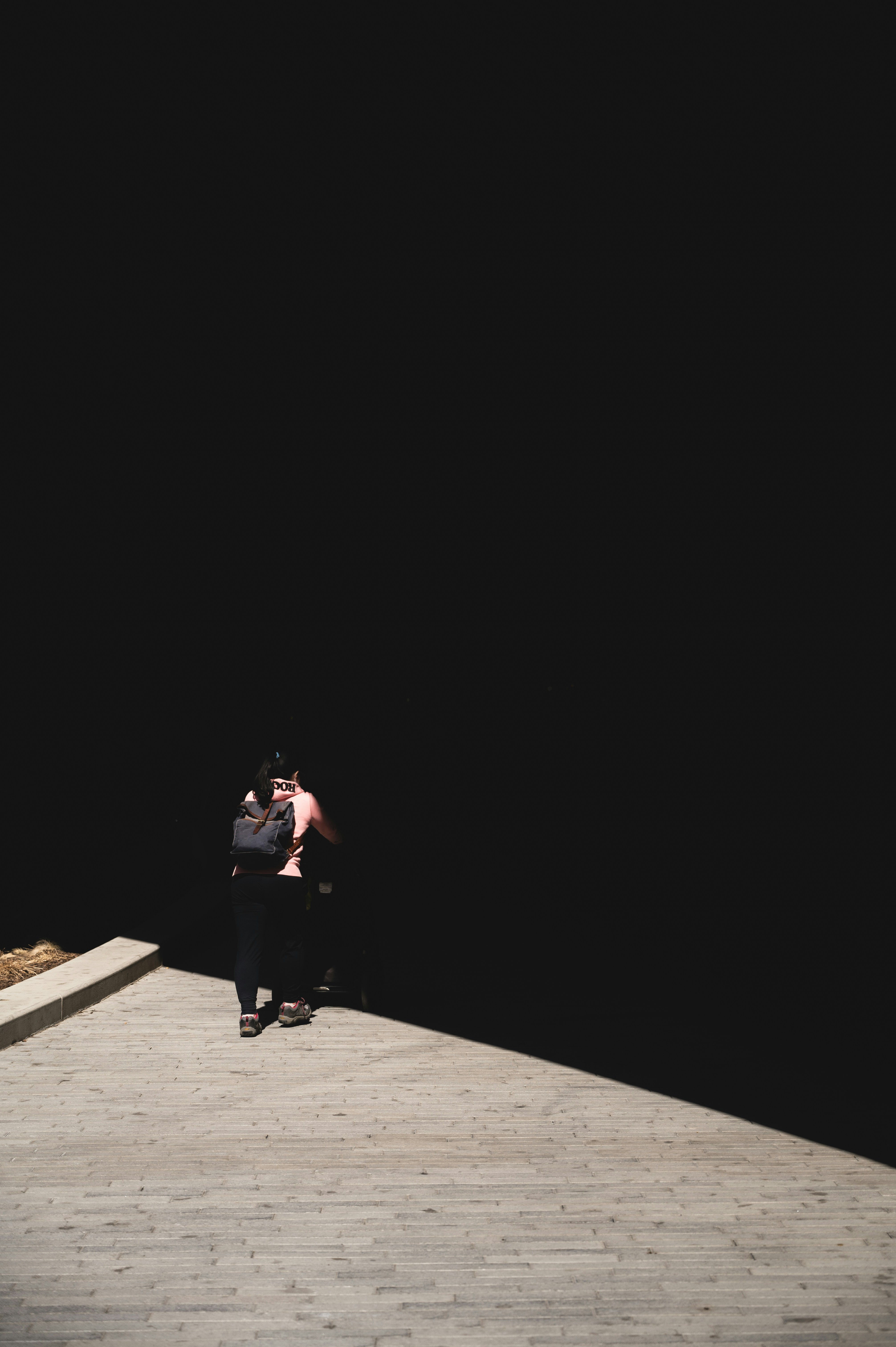 Two individuals walking along a wooden pathway leading into a dark expanse. The contrasting light and shadow create a sense of mystery.
