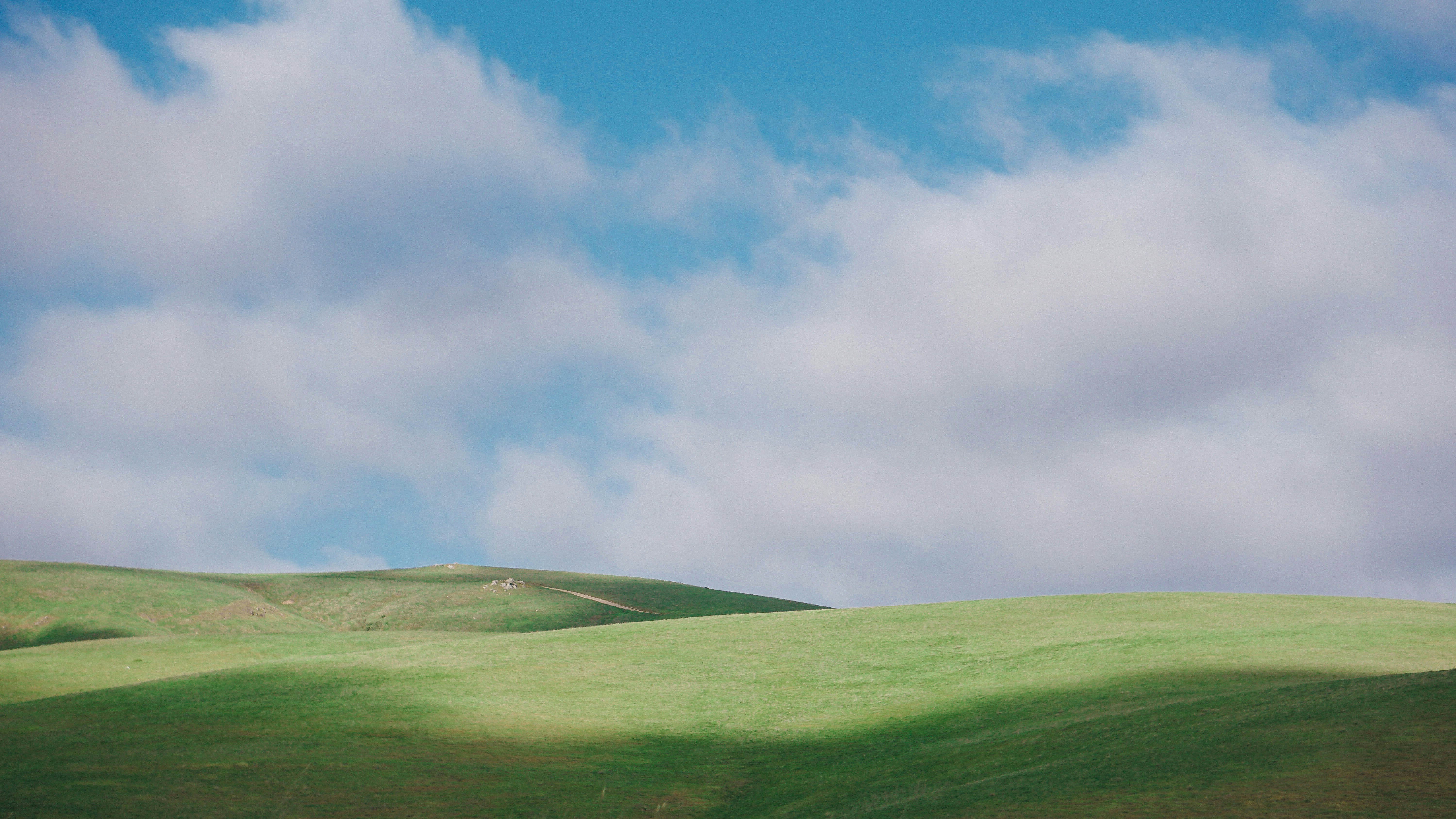 Vibrant green hills under a vast blue sky dotted with fluffy clouds. The scene conveys a serene, pastoral landscape.