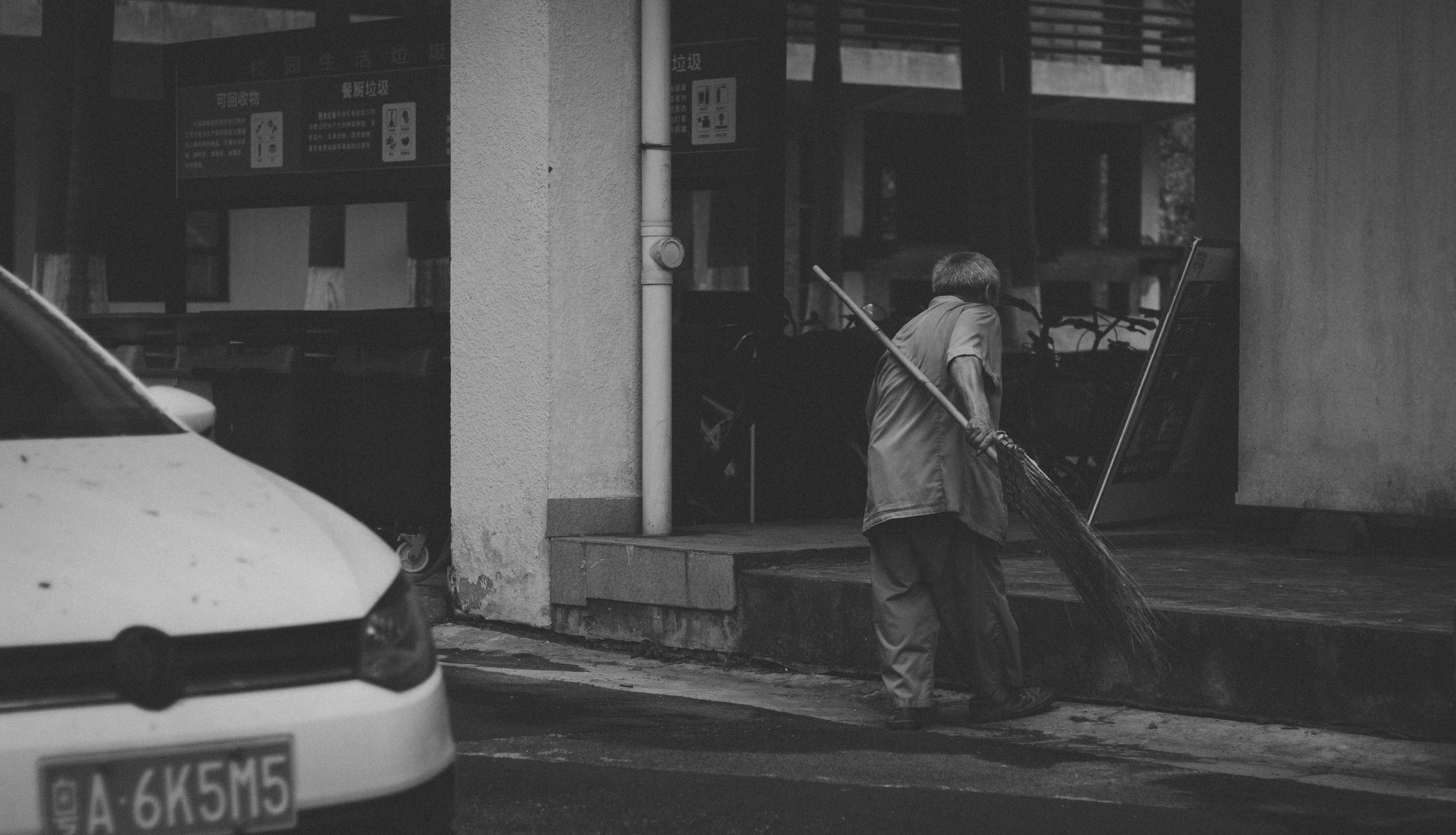 Elderly figure sweeping the pavement in an urban setting, exuding a sense of routine and diligence. Black and white tones enhance the mood.