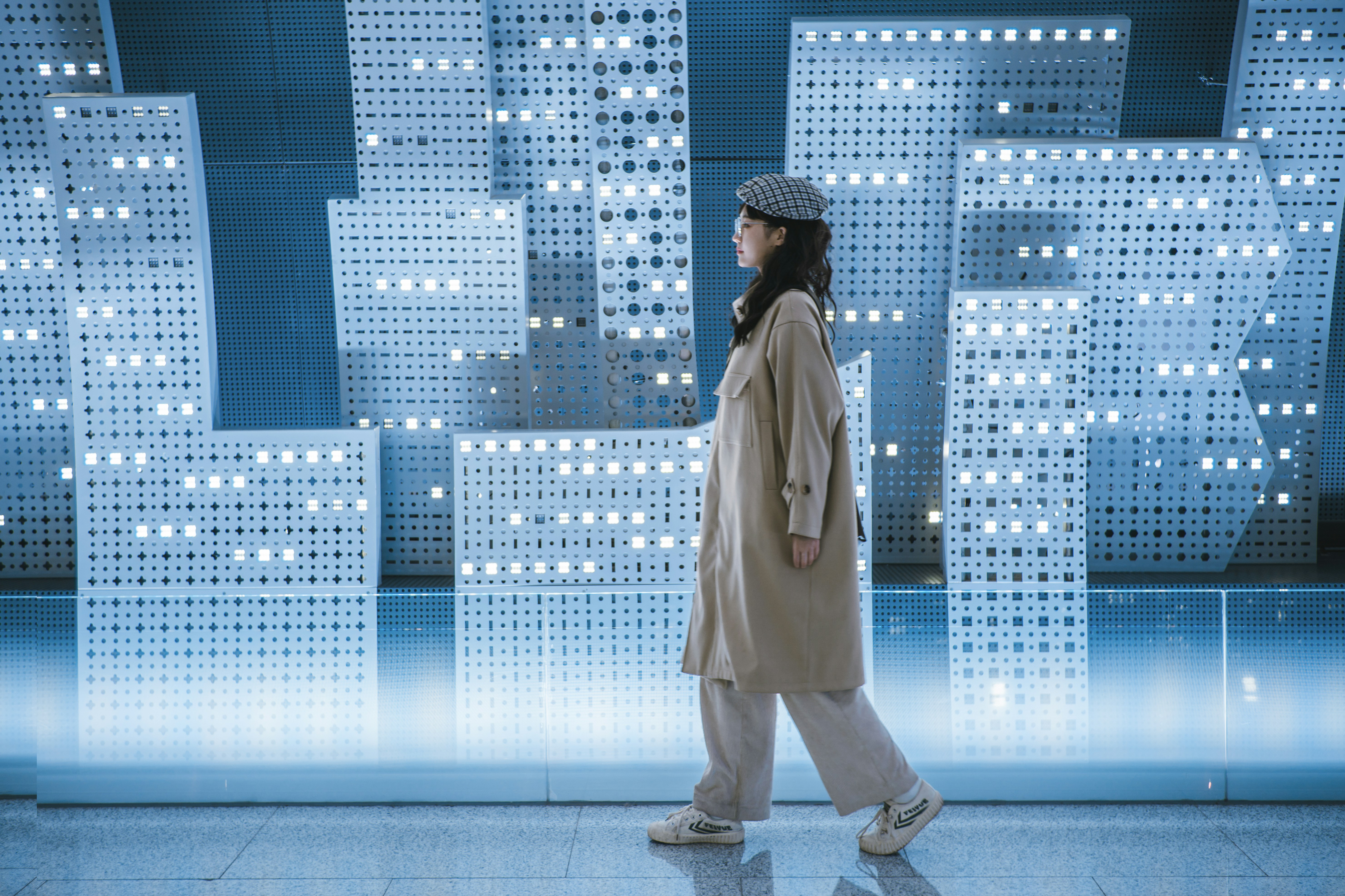 A woman in a stylish trench coat walks past a backdrop of illuminated, abstract cityscape panels. The scene blends modern fashion with innovative design.