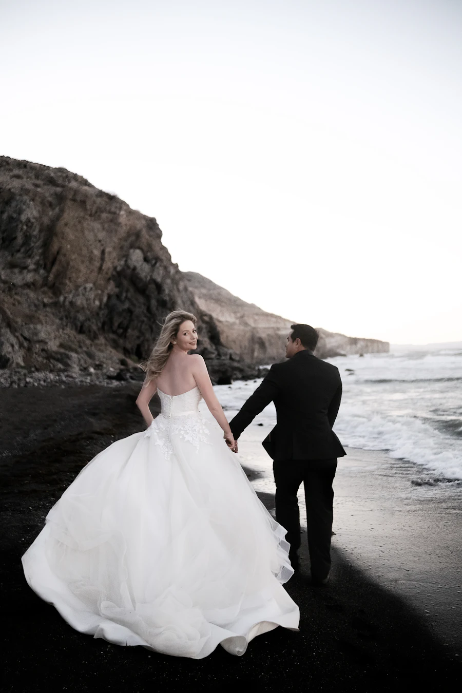 Bride in a flowing white gown and groom in a black suit holding hands while walking along a dark rocky beach beside the ocean.