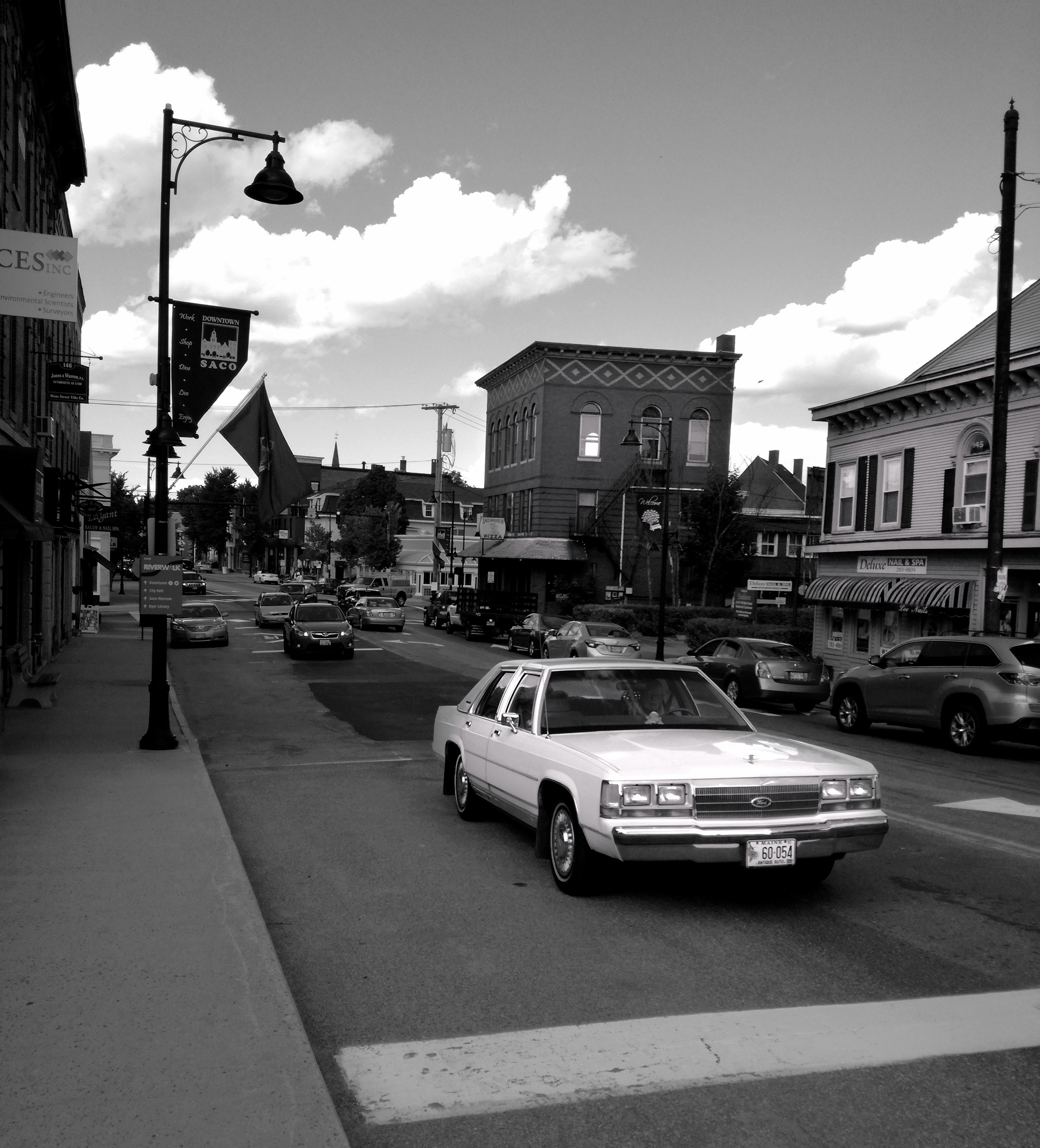 Classic American car cruising down a busy urban street lined with historic buildings under a partly cloudy sky.