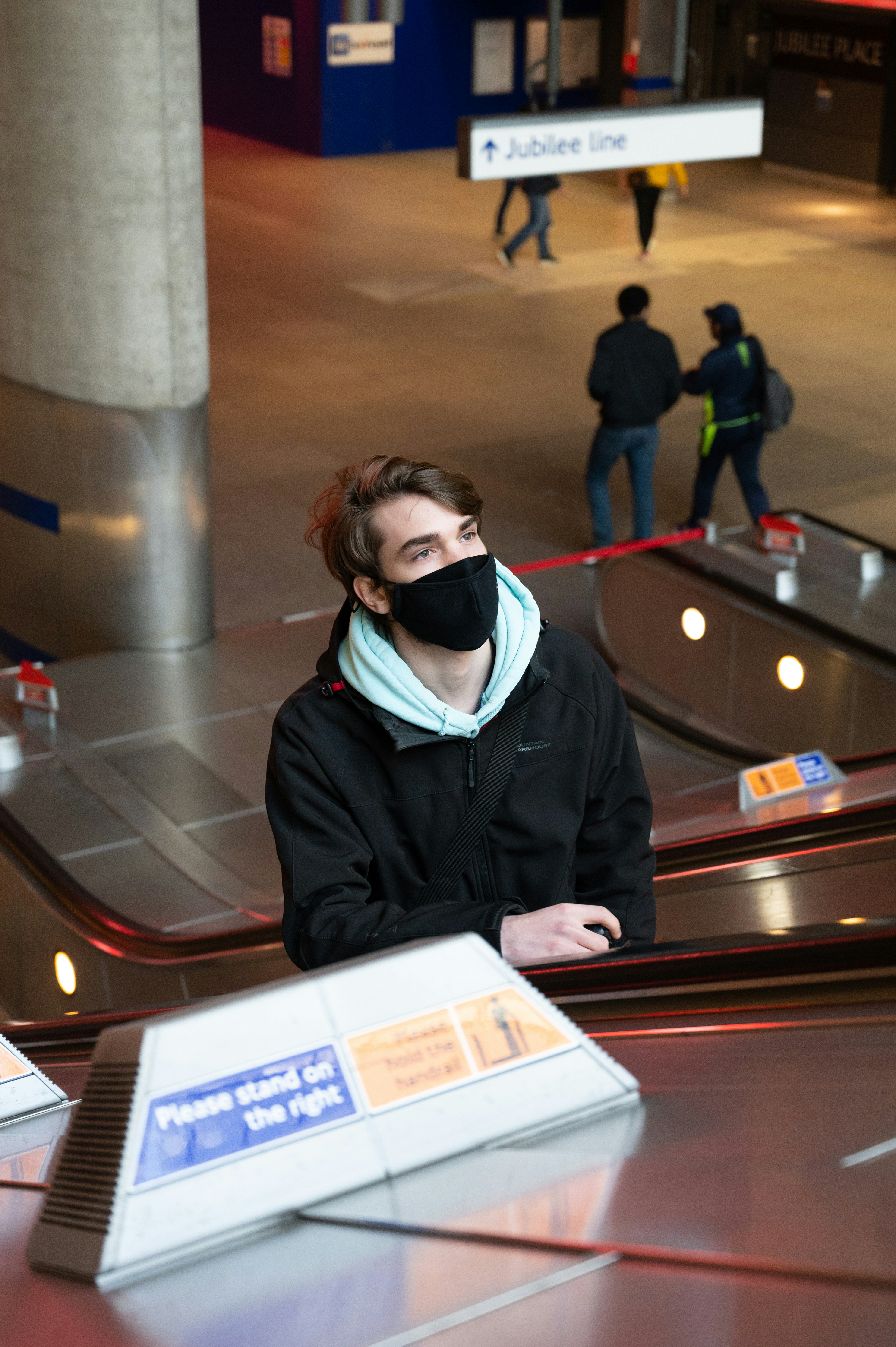 Young man on escalator in a transit station, wearing a mask and looking thoughtfully ahead. Signage indicates the Jubilee Line.