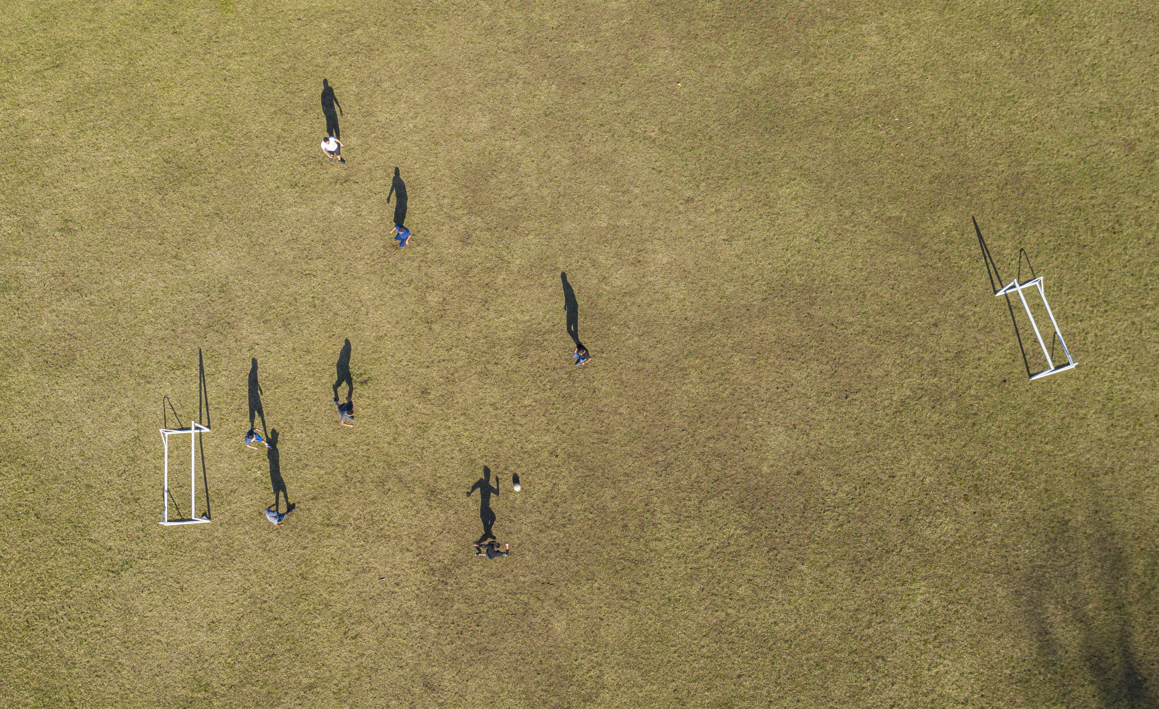 Aerial view of players practicing on a grass field, casting long shadows. Training equipment is scattered around, enhancing the scene's activity.