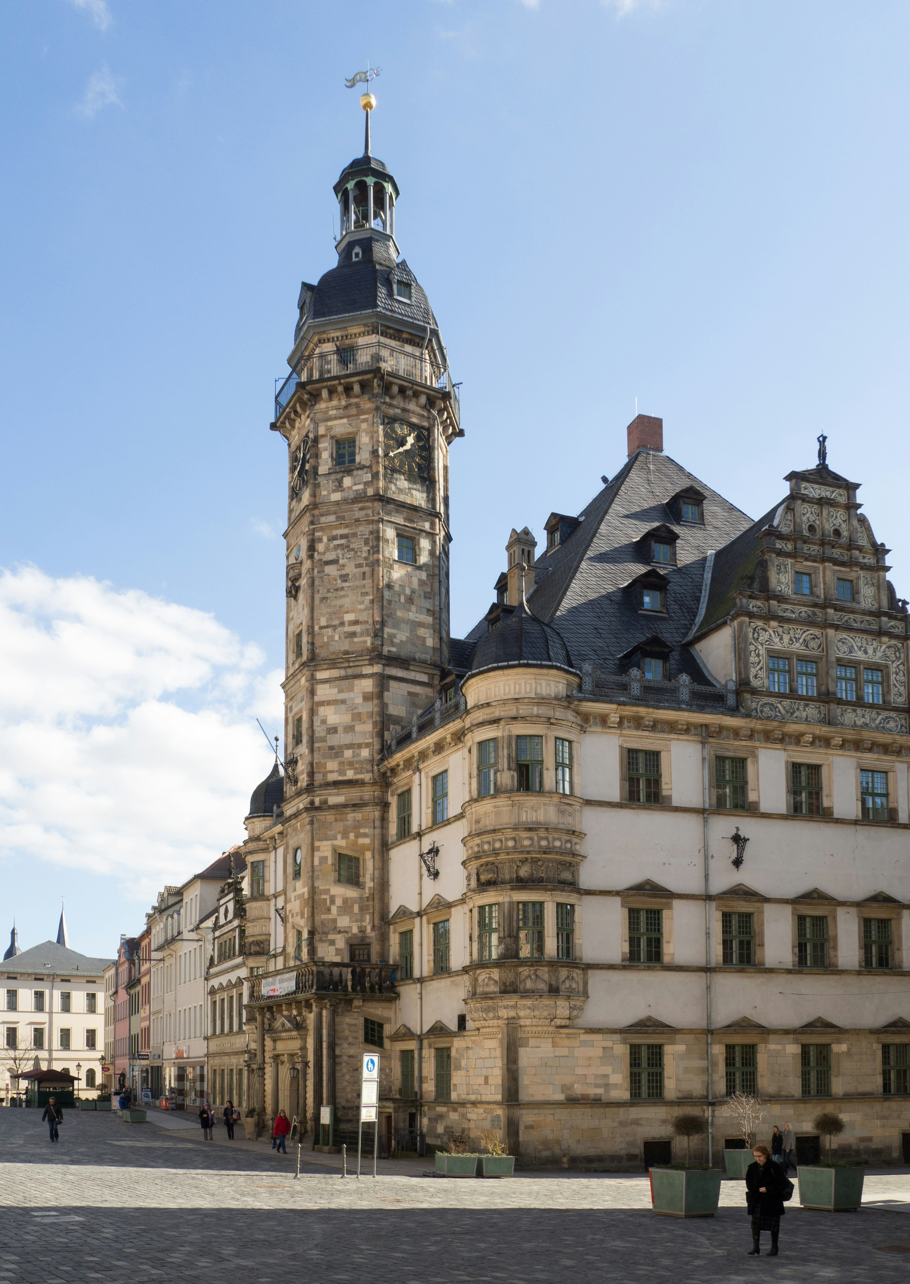 Historic building with a prominent clock tower and intricate architectural details, set against a clear blue sky. The scene captures the essence of a vibrant town square.