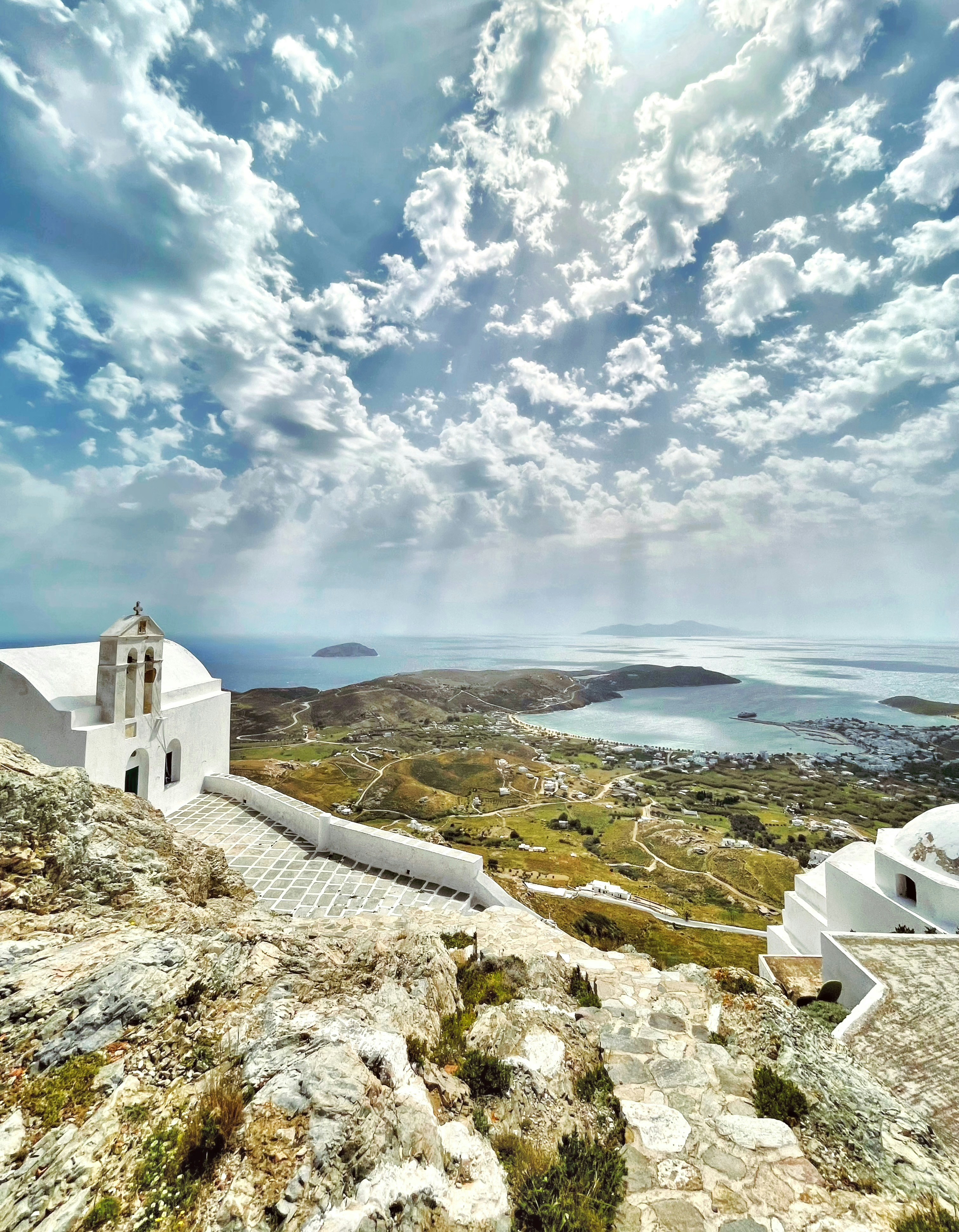 Whitewashed architecture perched on rocky terrain, overlooking a serene Aegean coastline under a dramatic sky.