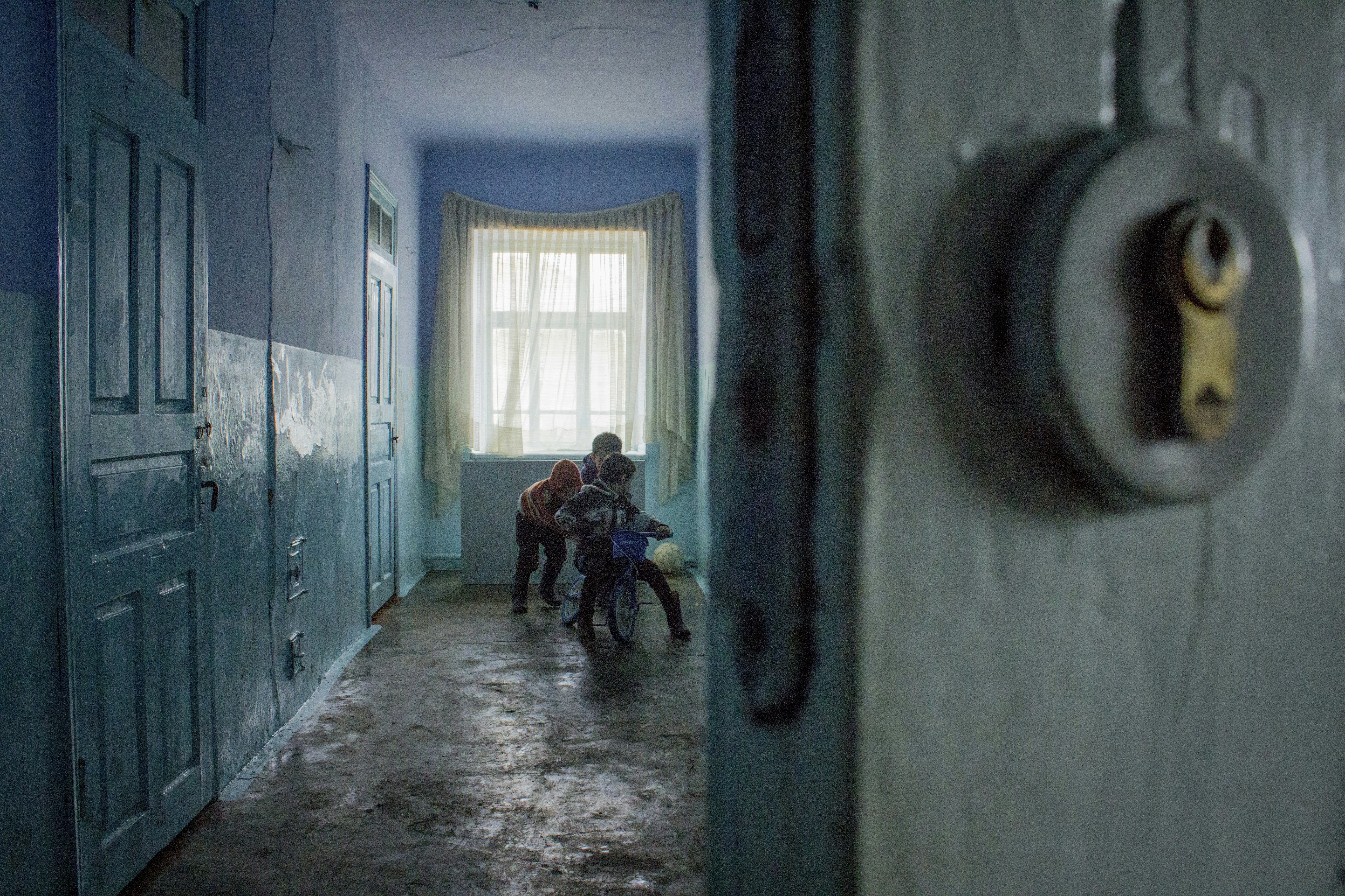 Two children playing on a bicycle in a dimly lit, worn hallway with peeling paint and a large window. The scene captures the essence of innocence amidst decay.