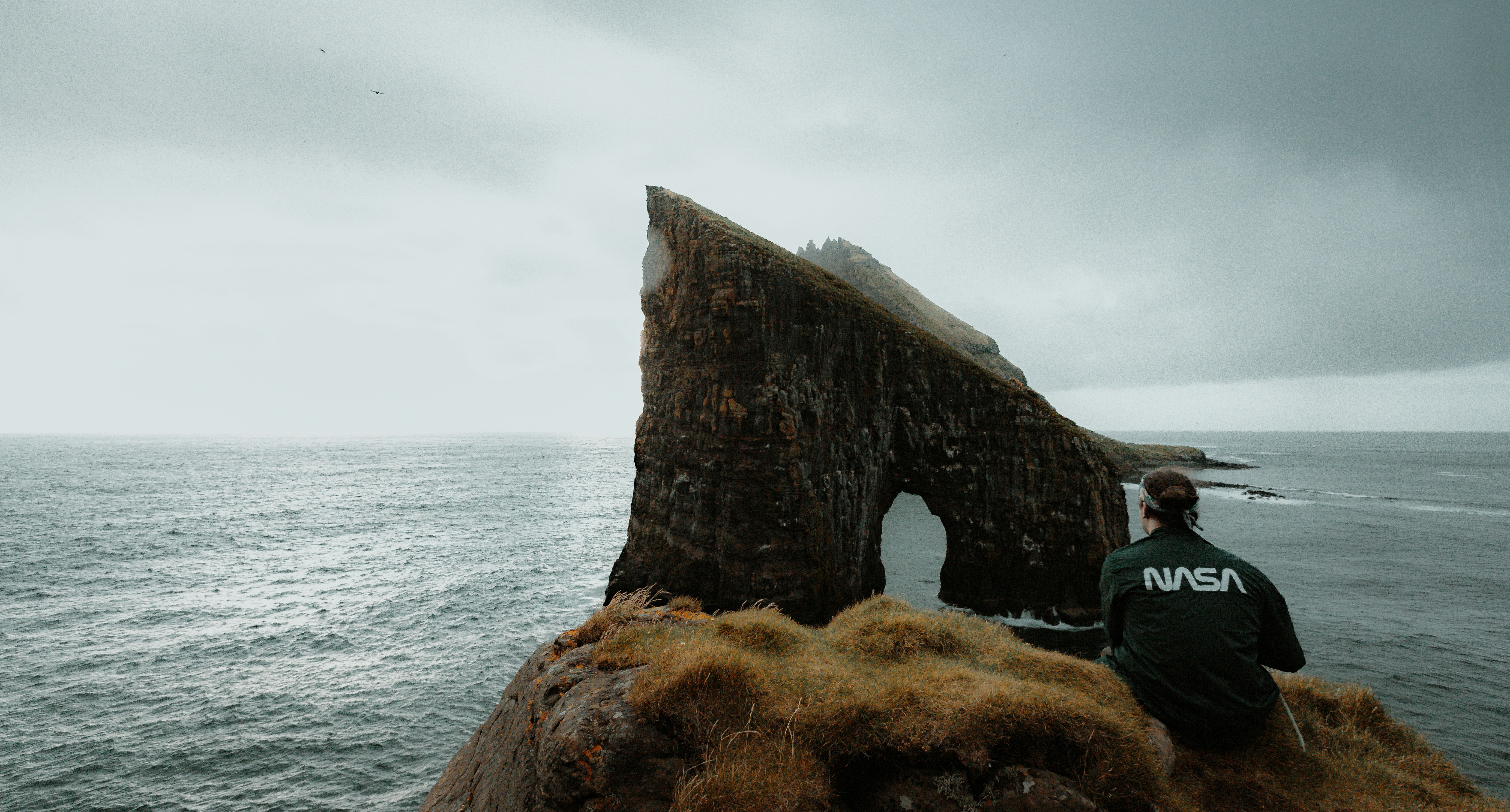 Solitary figure gazes at a rugged sea arch jutting from the ocean under a moody sky.