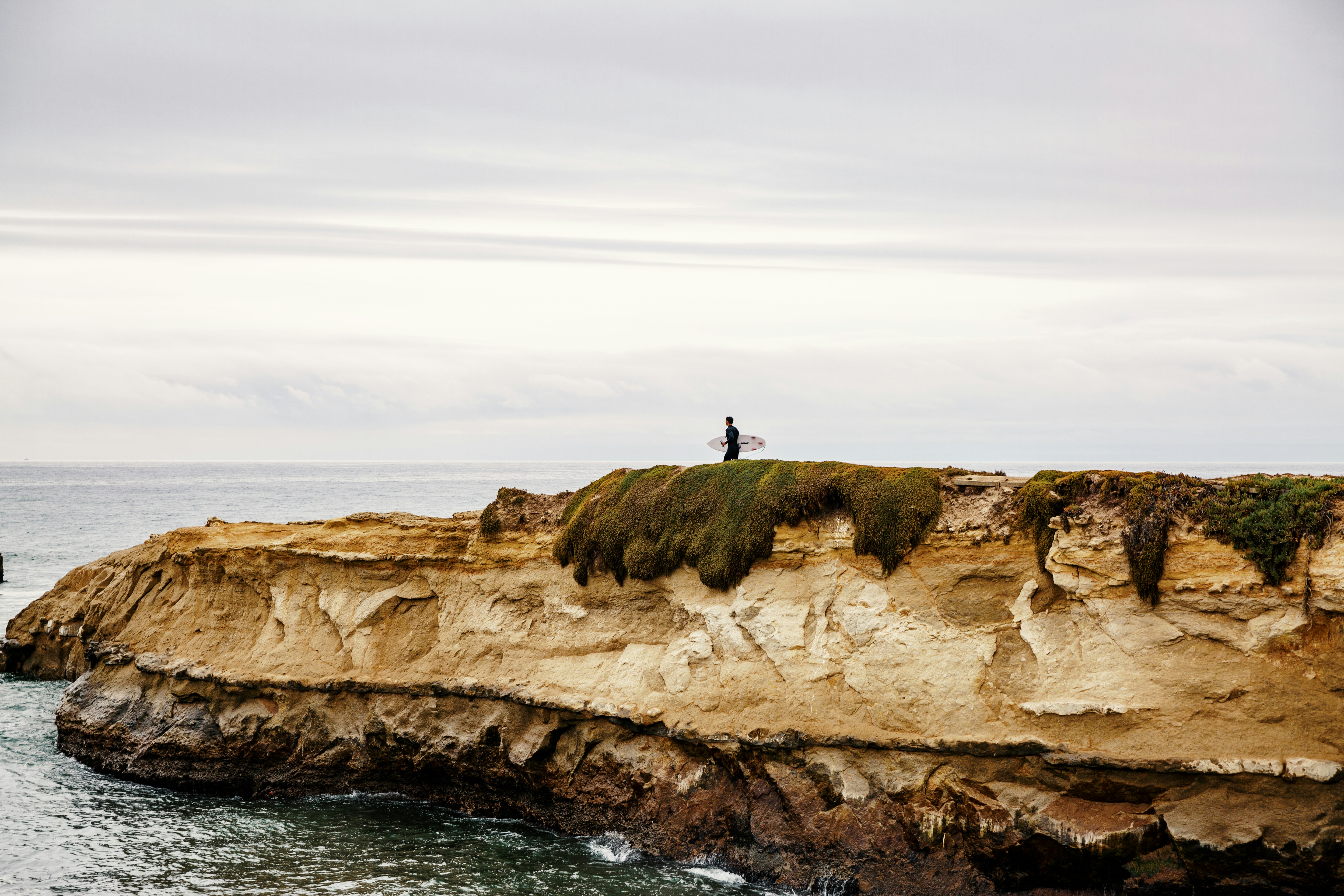 Person standing atop rugged cliff overlooking ocean under overcast sky.