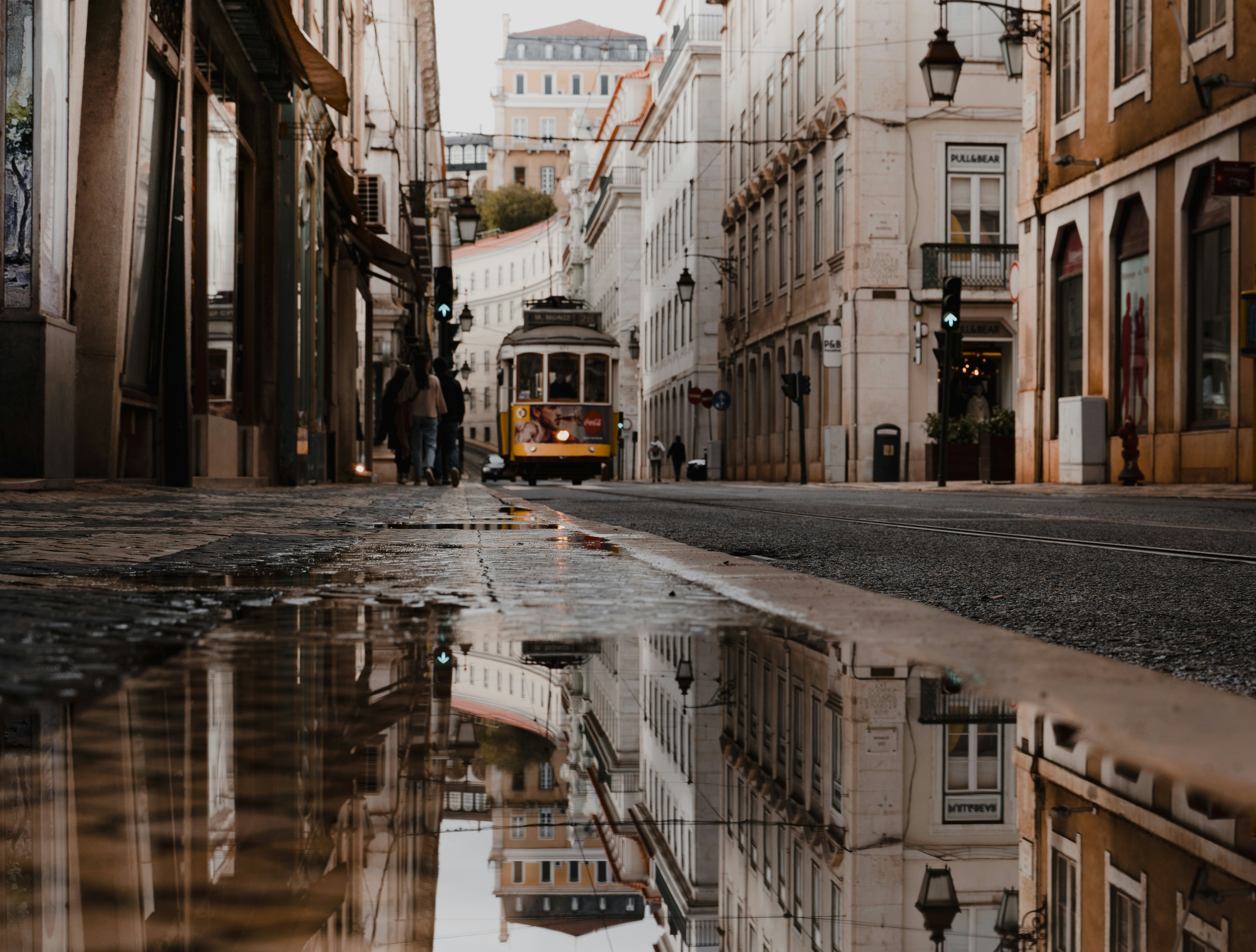 Vintage tram gliding through a narrow Lisbon street, reflected in a puddle after rain, showcasing the city's charming architecture.