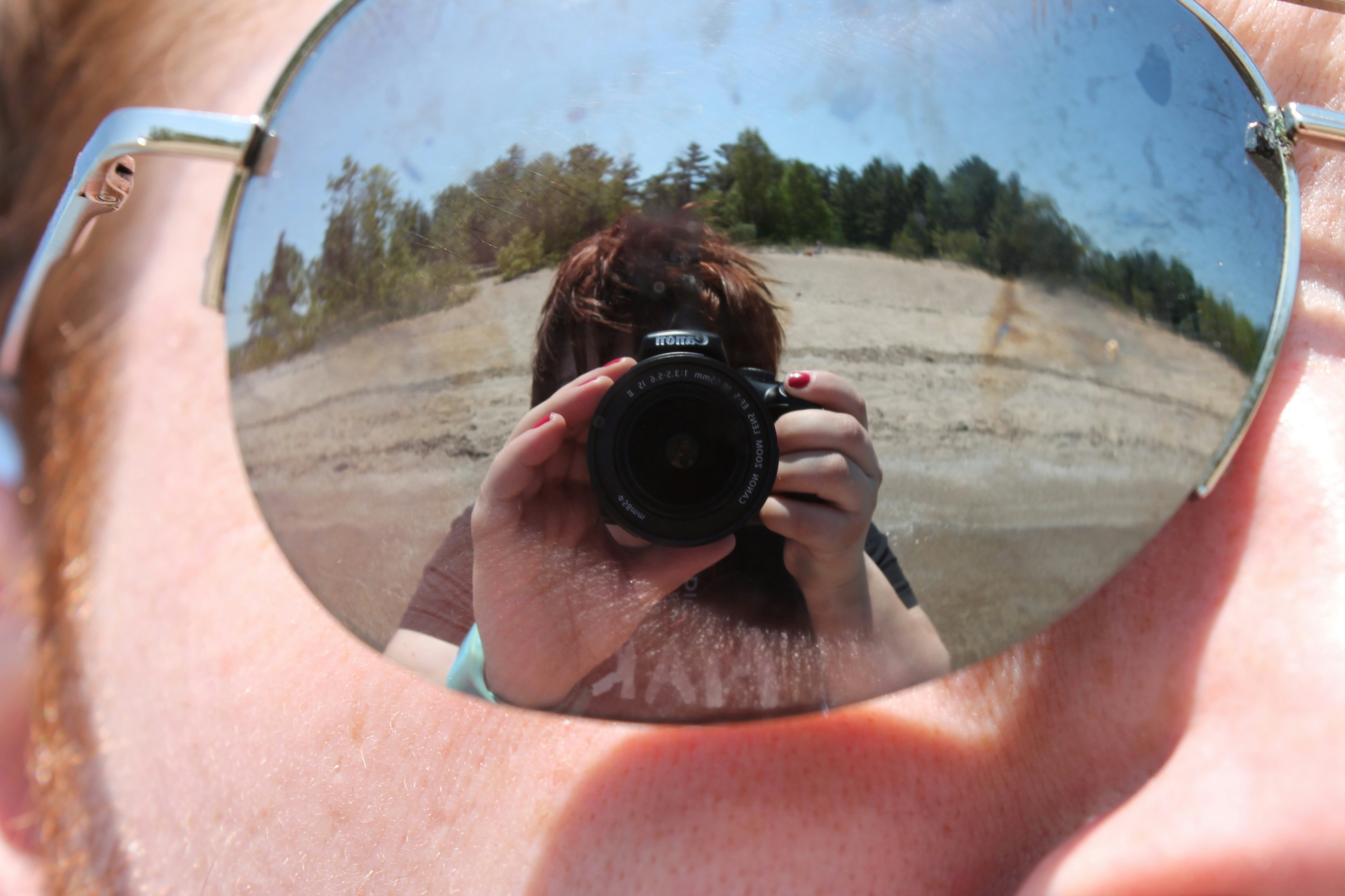 Self-portrait in reflective sunglasses shows the photographer holding a camera, with a sunlit landscape mirrored in the lenses.
