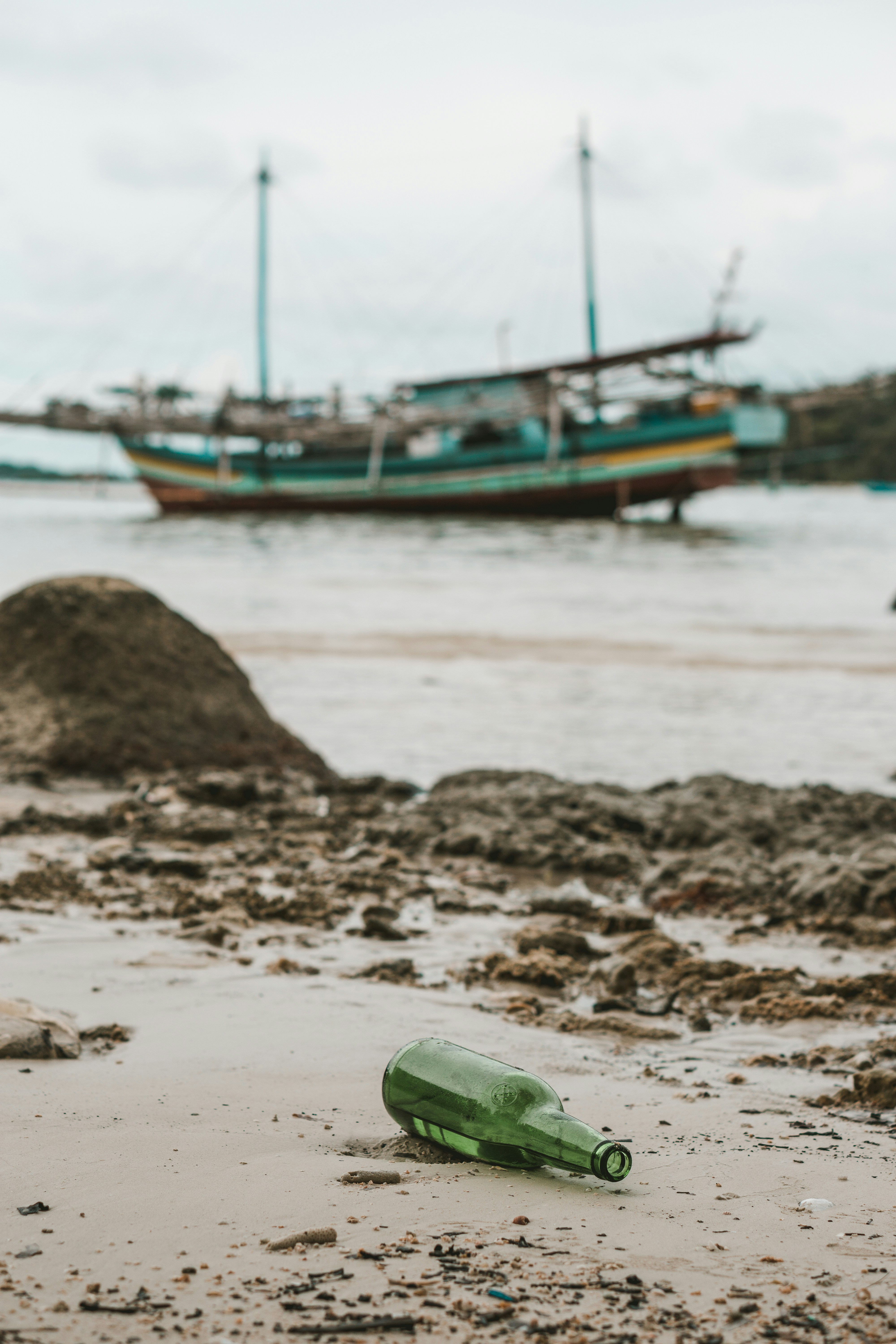 A discarded green bottle rests on a sandy beach, with a traditional boat anchored in the background, hinting at the intersection of nature and human impact.