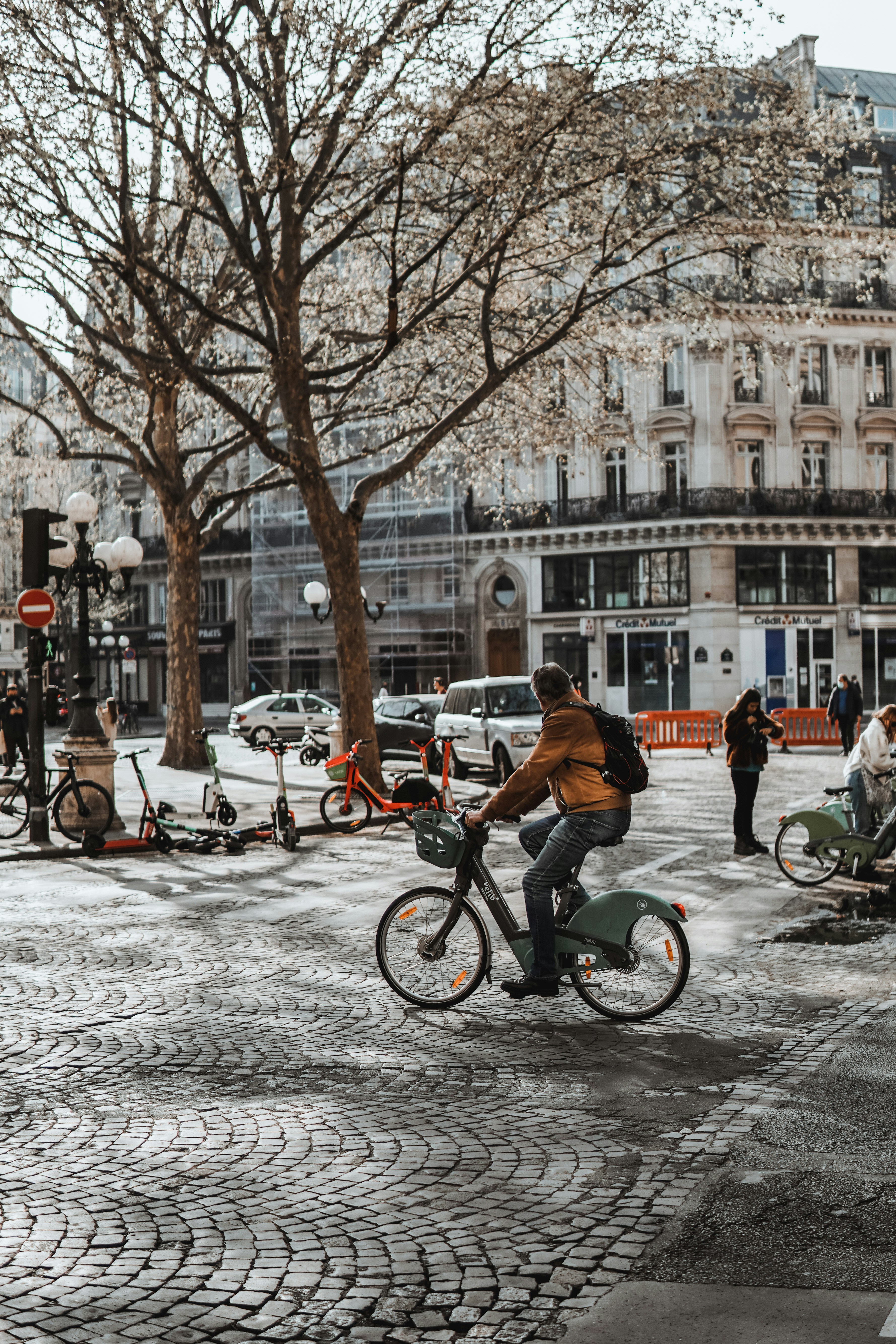 A cyclist navigates a bustling urban intersection lined with trees and parked bikes. The scene captures the essence of city life and mobility.