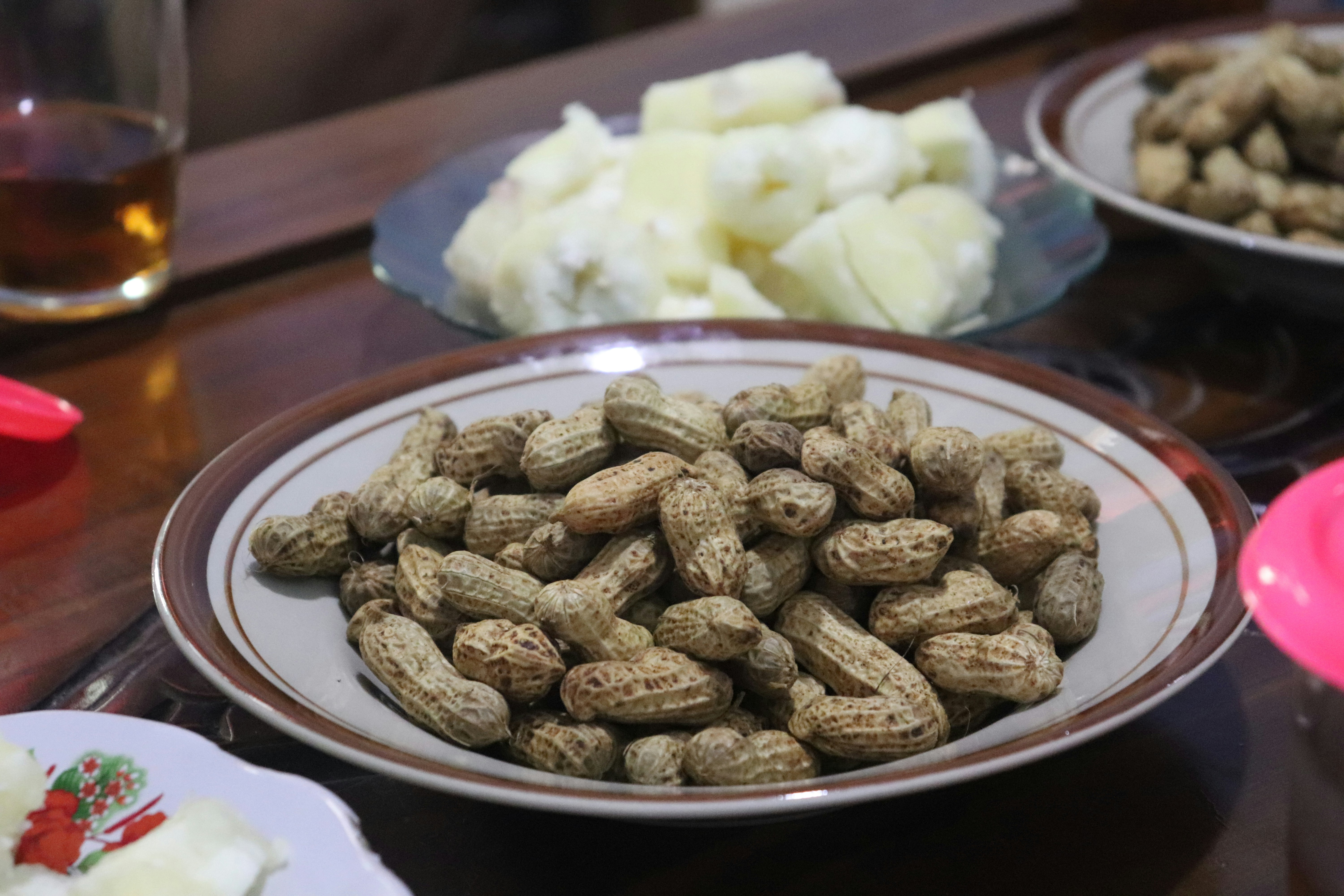 Boiled peanuts in a ceramic bowl surrounded by sliced yams and a glass of tea on a wooden table.