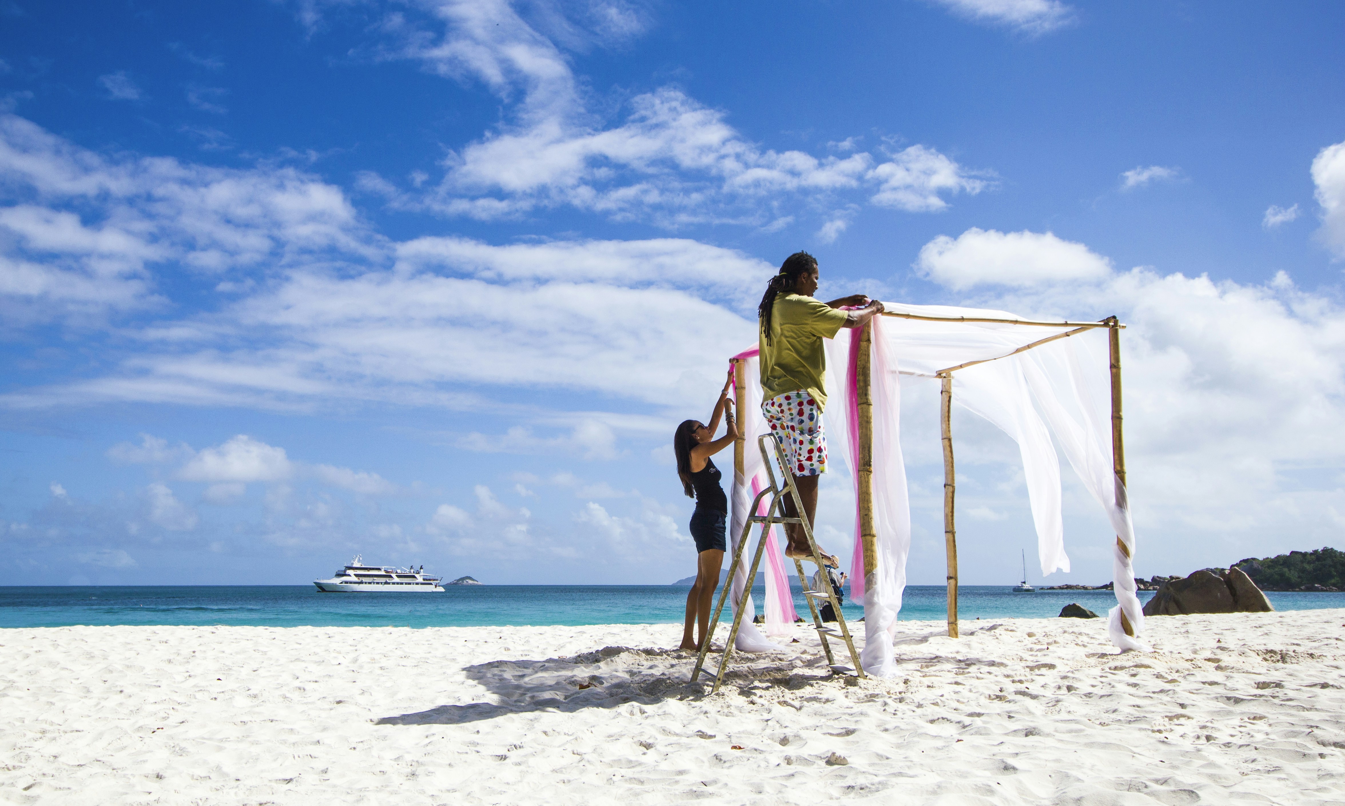 Two individuals arranging flowing fabrics on a wooden structure at a picturesque beach, with a boat in the background. The vibrant colors contrast beautifully against the serene ocean.