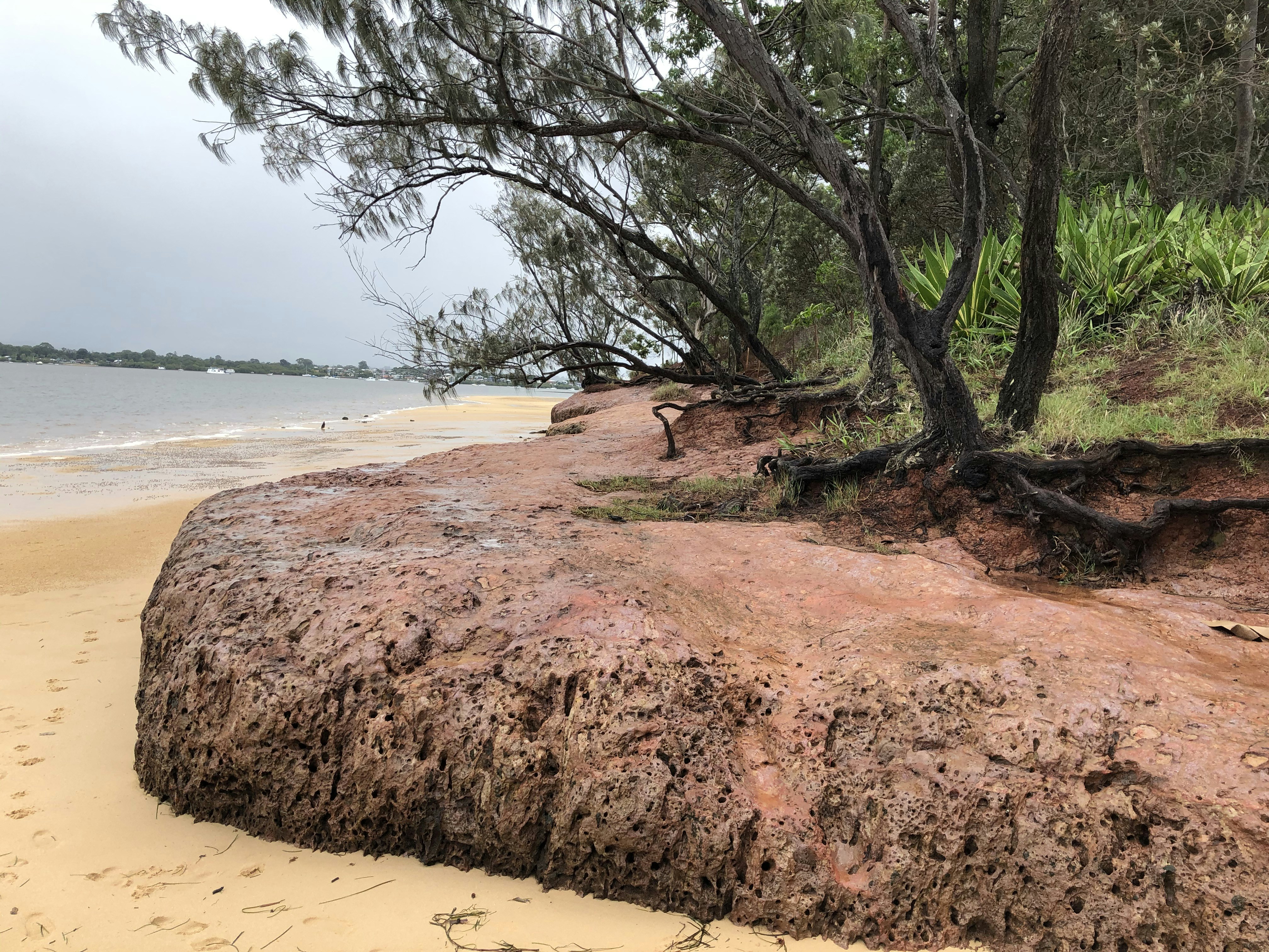 Rugged red rock formations meet the sandy beach under a cloudy sky, with trees framing the scene on the edge of the water.