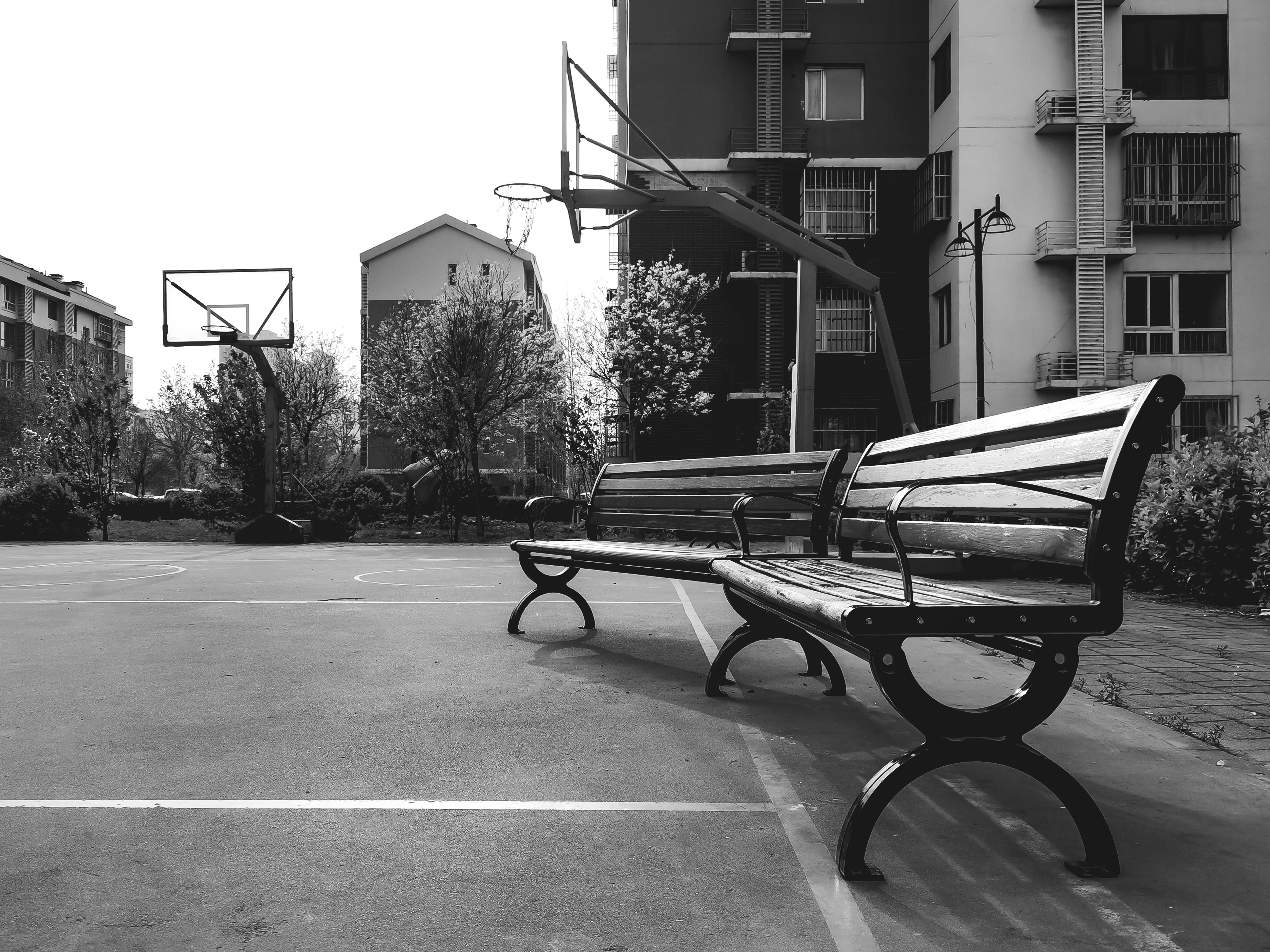 Black-and-white photograph of a lone bench along a basketball court, with a distant hoop and urban buildings in the background. The composition centers the bench as a quiet anchor in the scene.