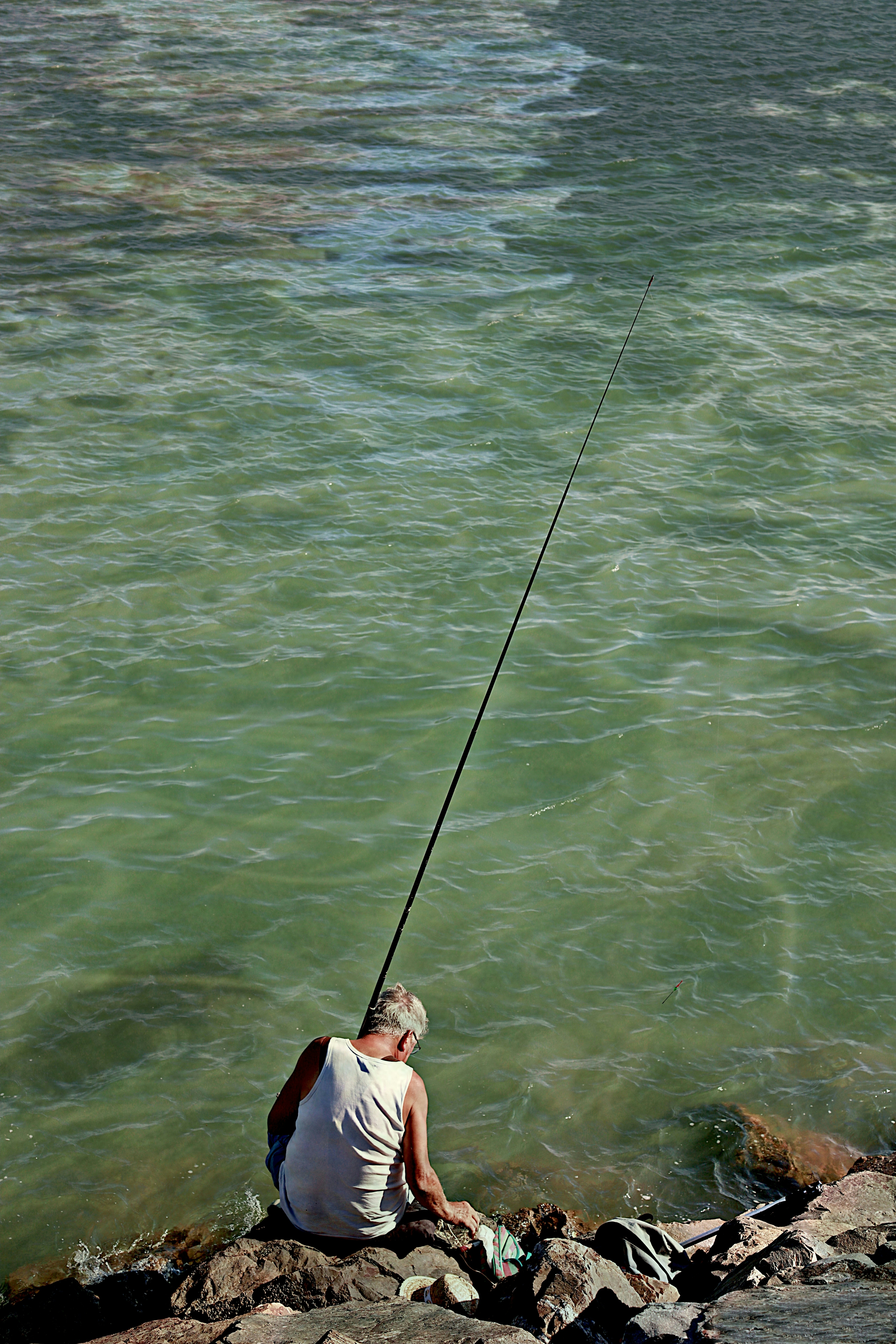 An elderly man fishing from rocky shores, focused on his catch while gentle waves lap at the stones. The tranquil water reflects the sky's hues.