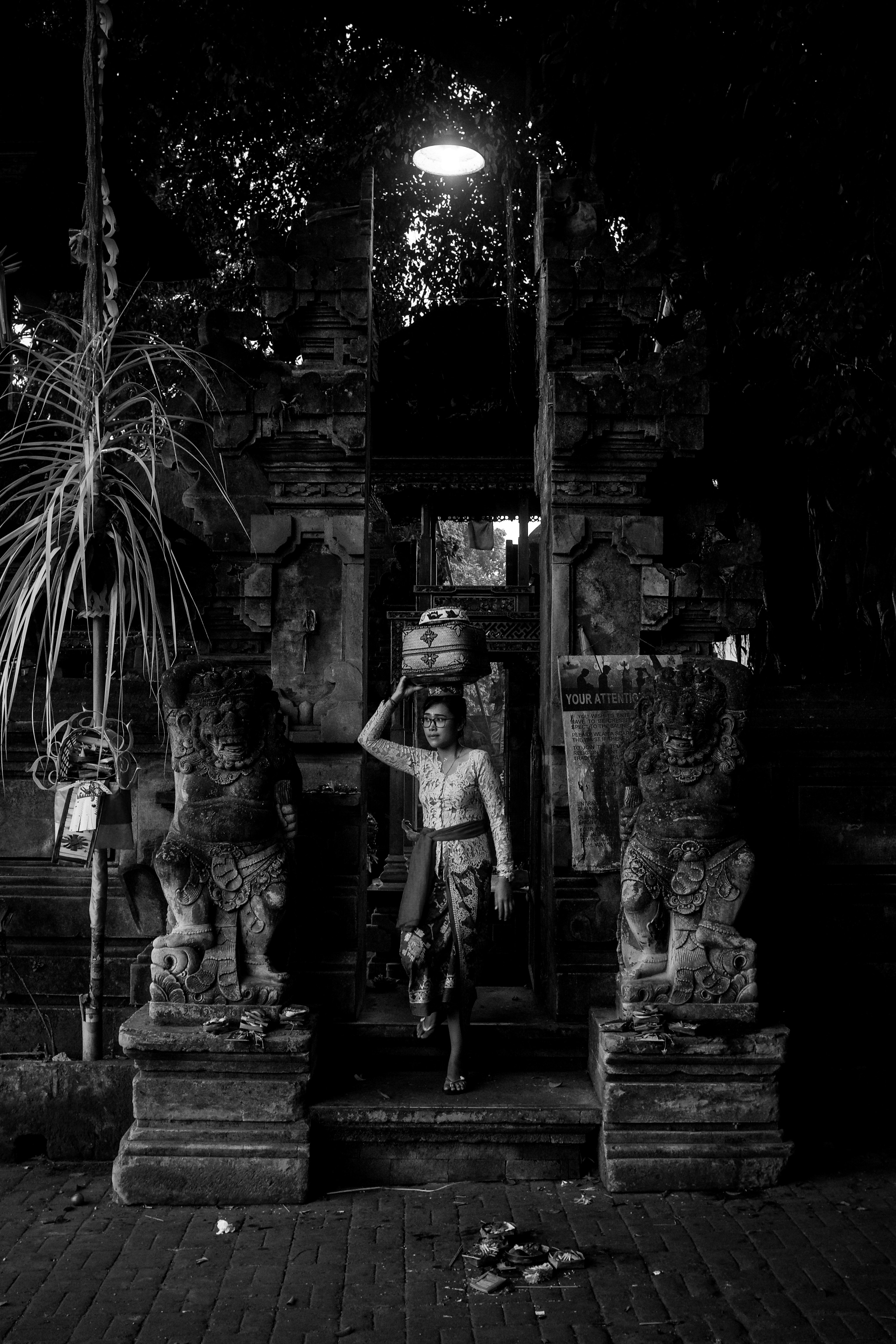A woman balancing a basket on her head walks past intricately carved stone guardians at a temple entrance, embodying cultural heritage.