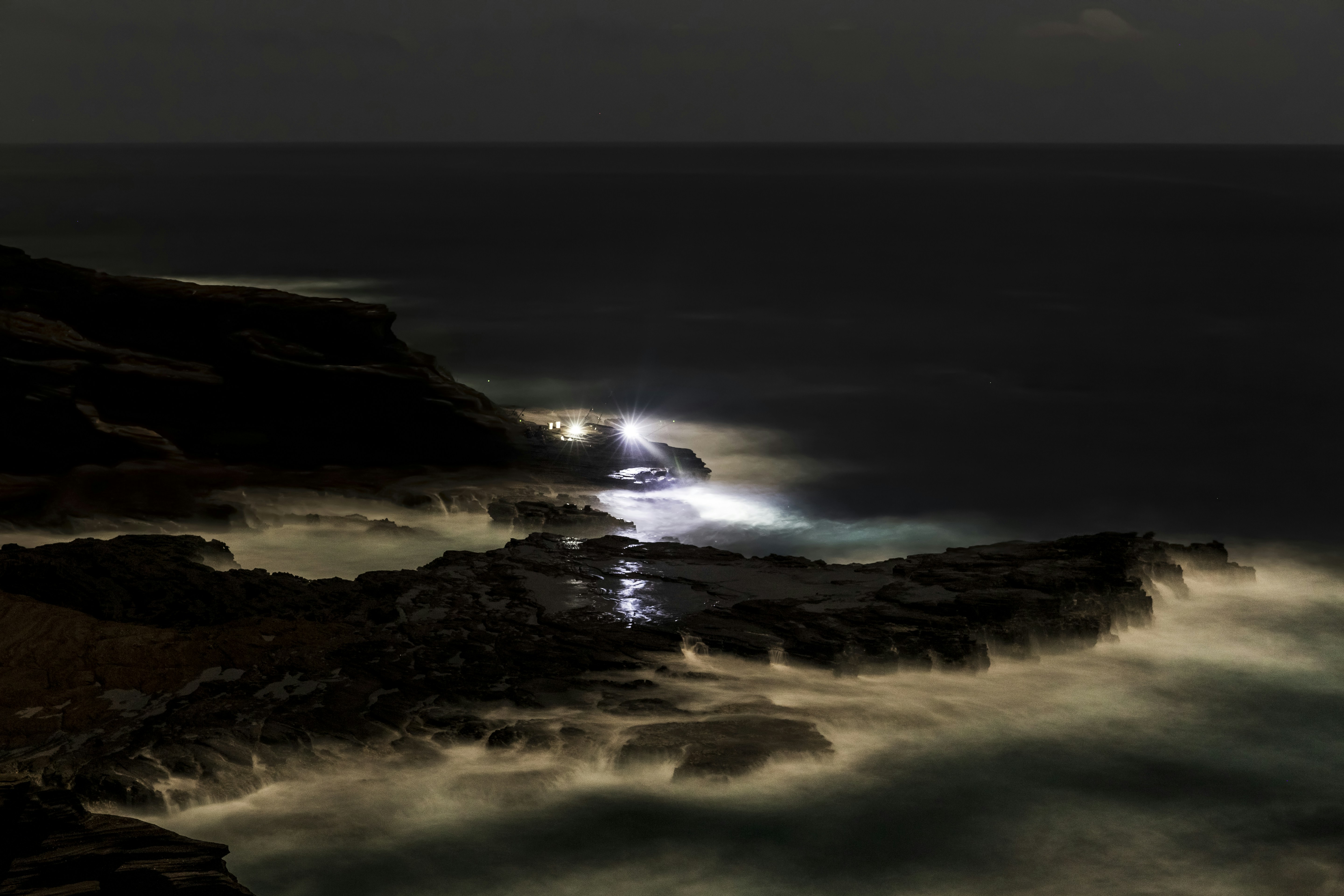 Moonlit rocks along a rugged coastline with gentle waves lapping at the shore, illuminated by distant lights. A serene nocturnal landscape.