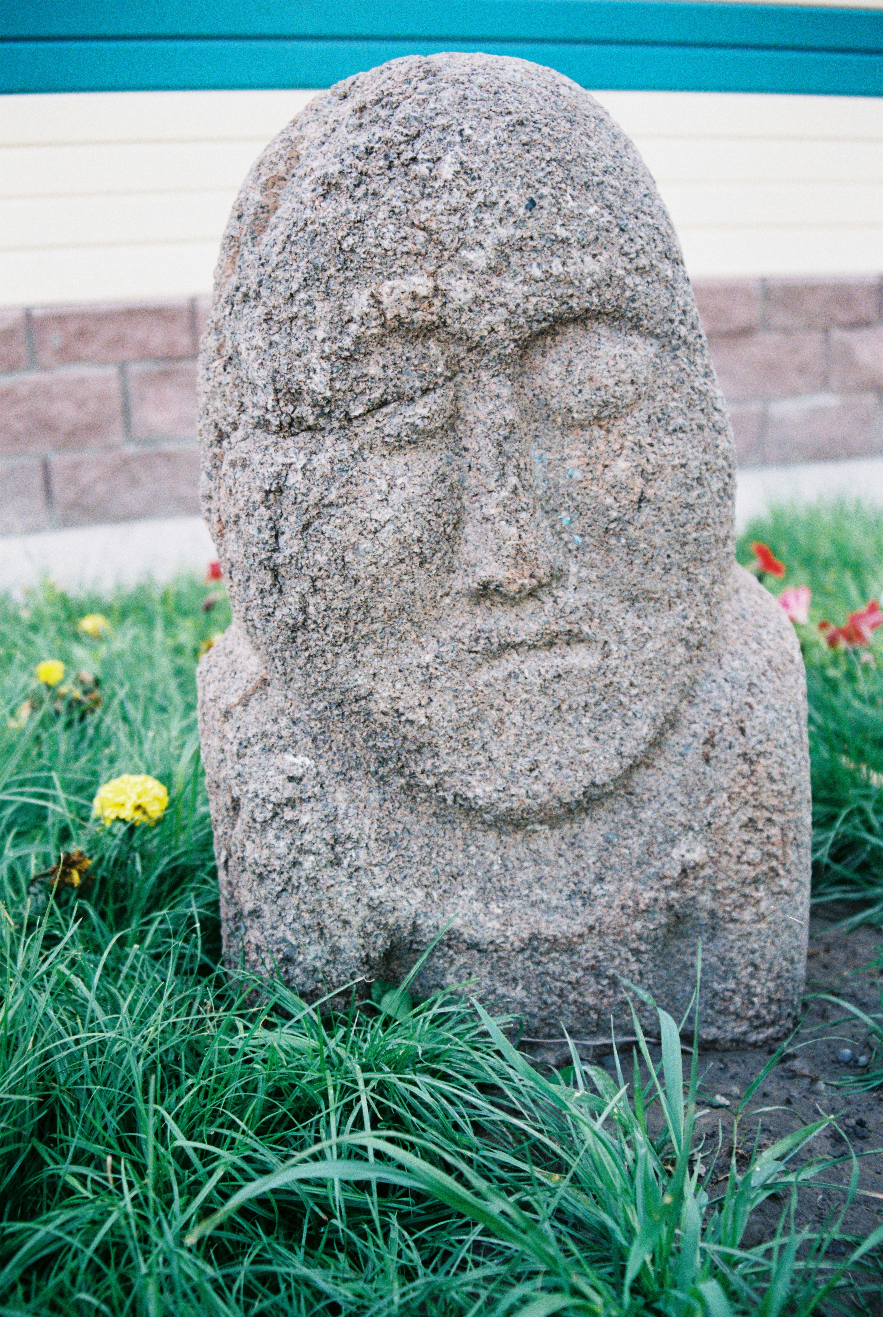 Granite sculpture of a serene face partially obscured by grass and flowers, evoking a sense of tranquility and history.