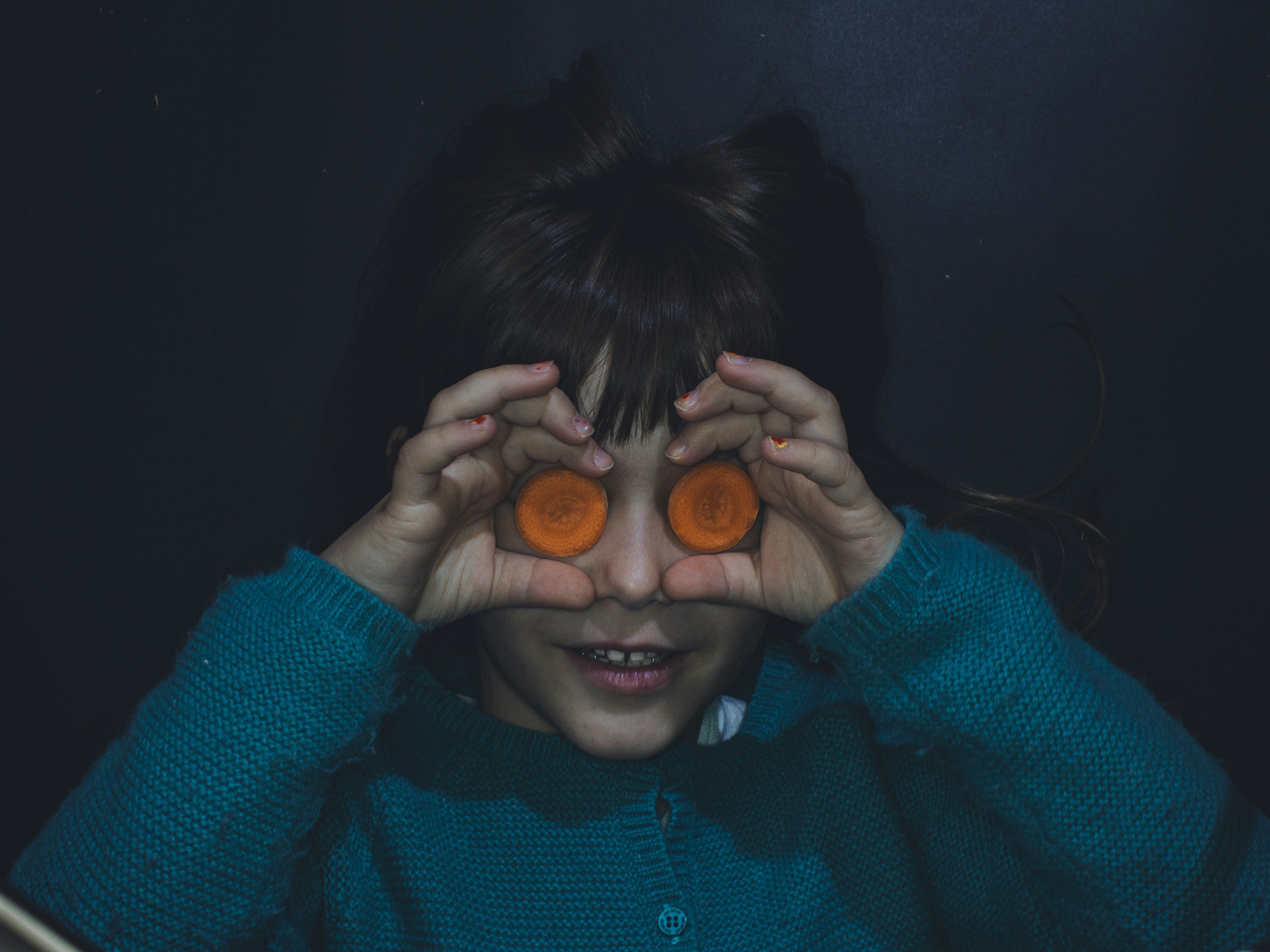 Child playfully holds carrot slices over their eyes, wearing a teal sweater against a dark background.