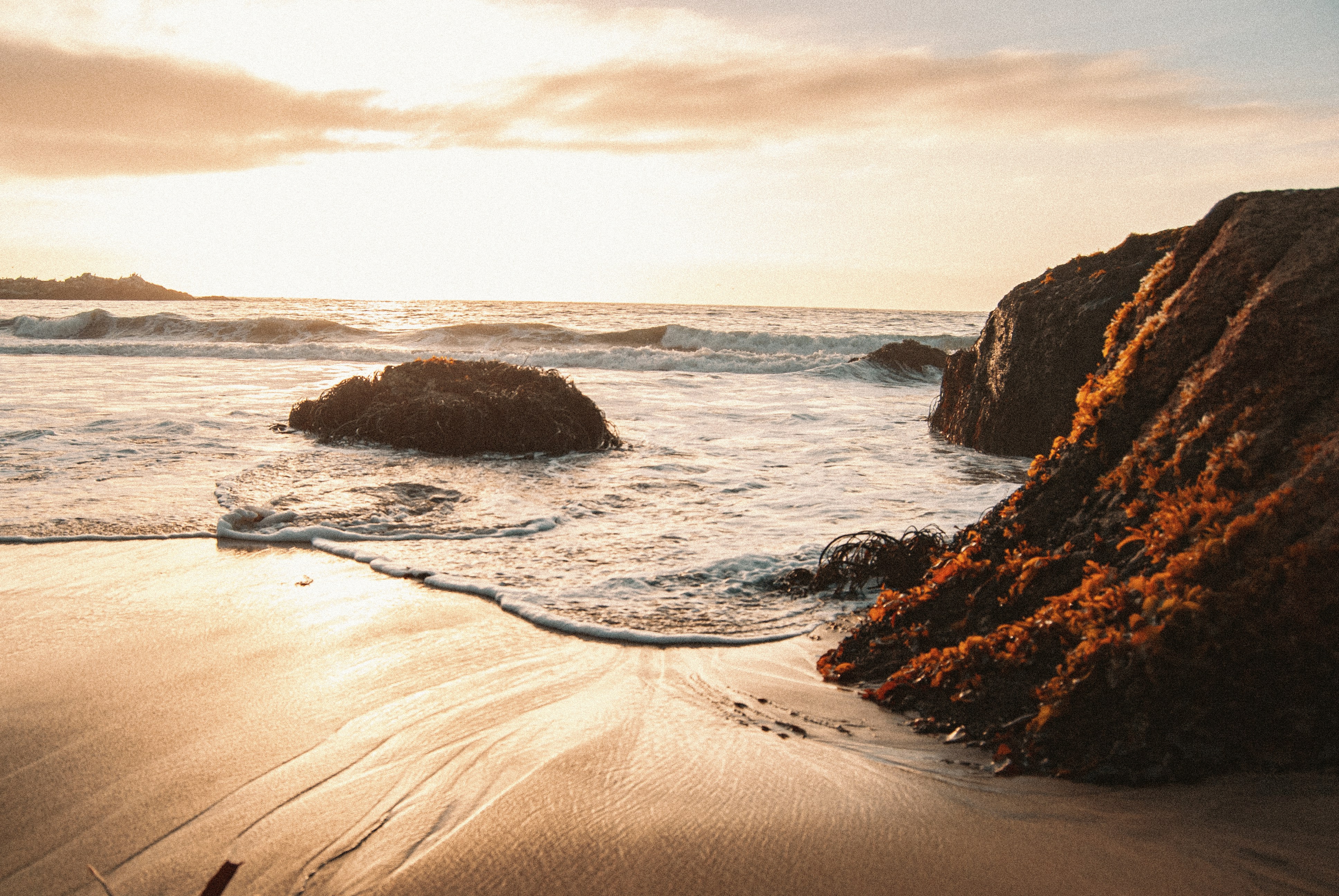 Sunset over a rocky beach with gentle waves and warm golden light reflecting off wet sand.