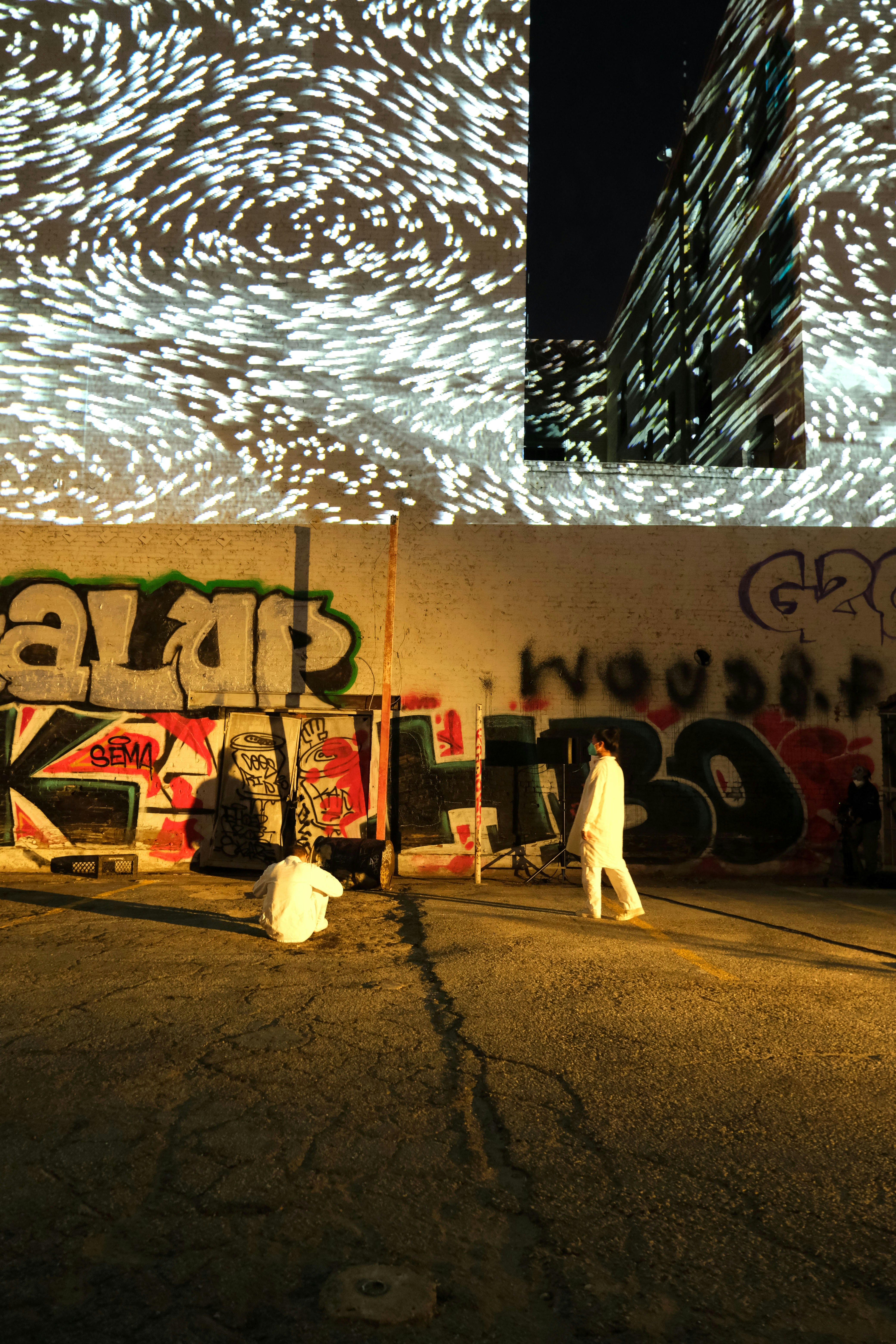 Two performers in white costumes interact with vibrant street art illuminated by a dynamic light projection. The scene captures the fusion of urban culture and artistic expression.