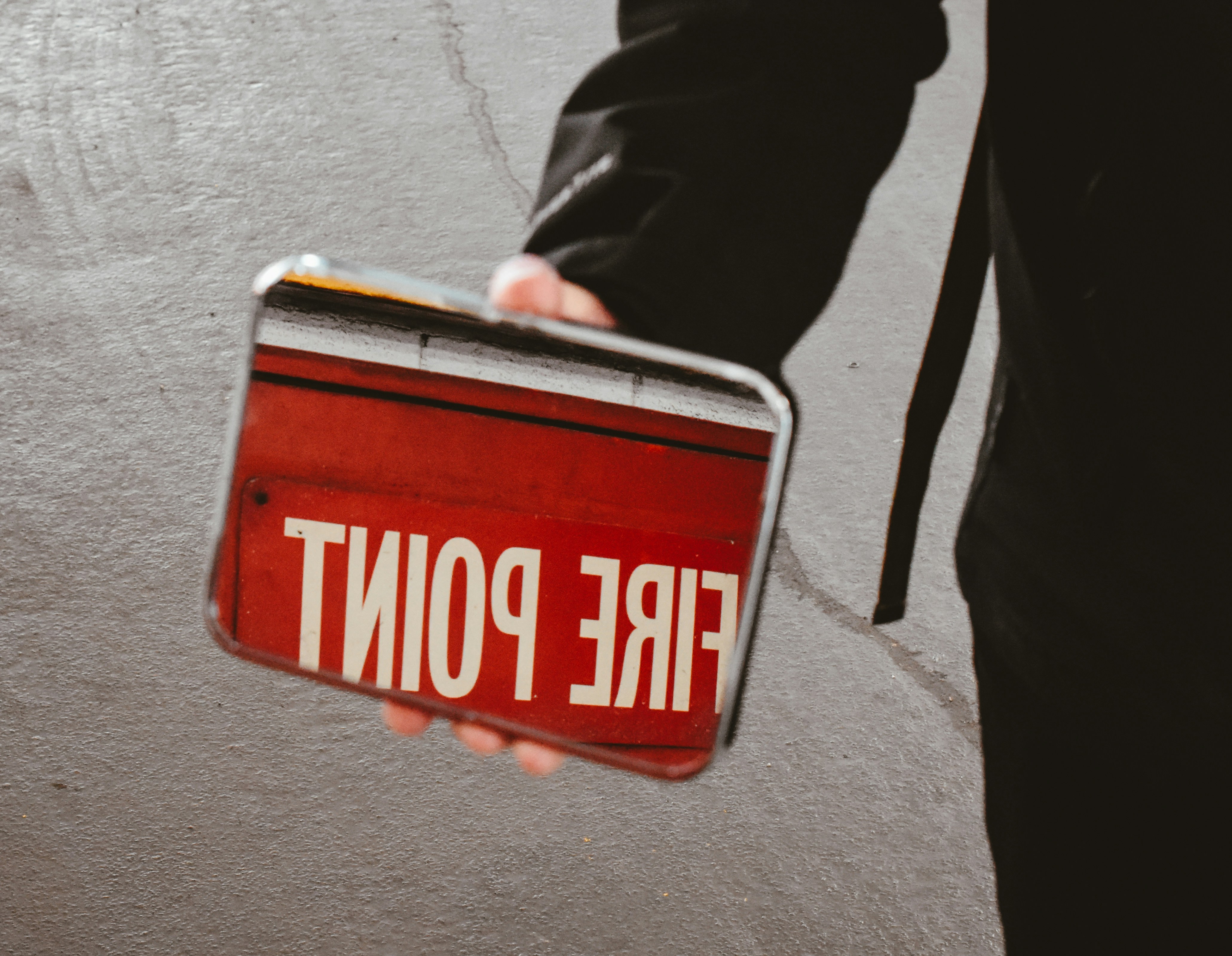 Red 'Fire Point' sign reflected upside-down in a handheld mirror against a concrete background.