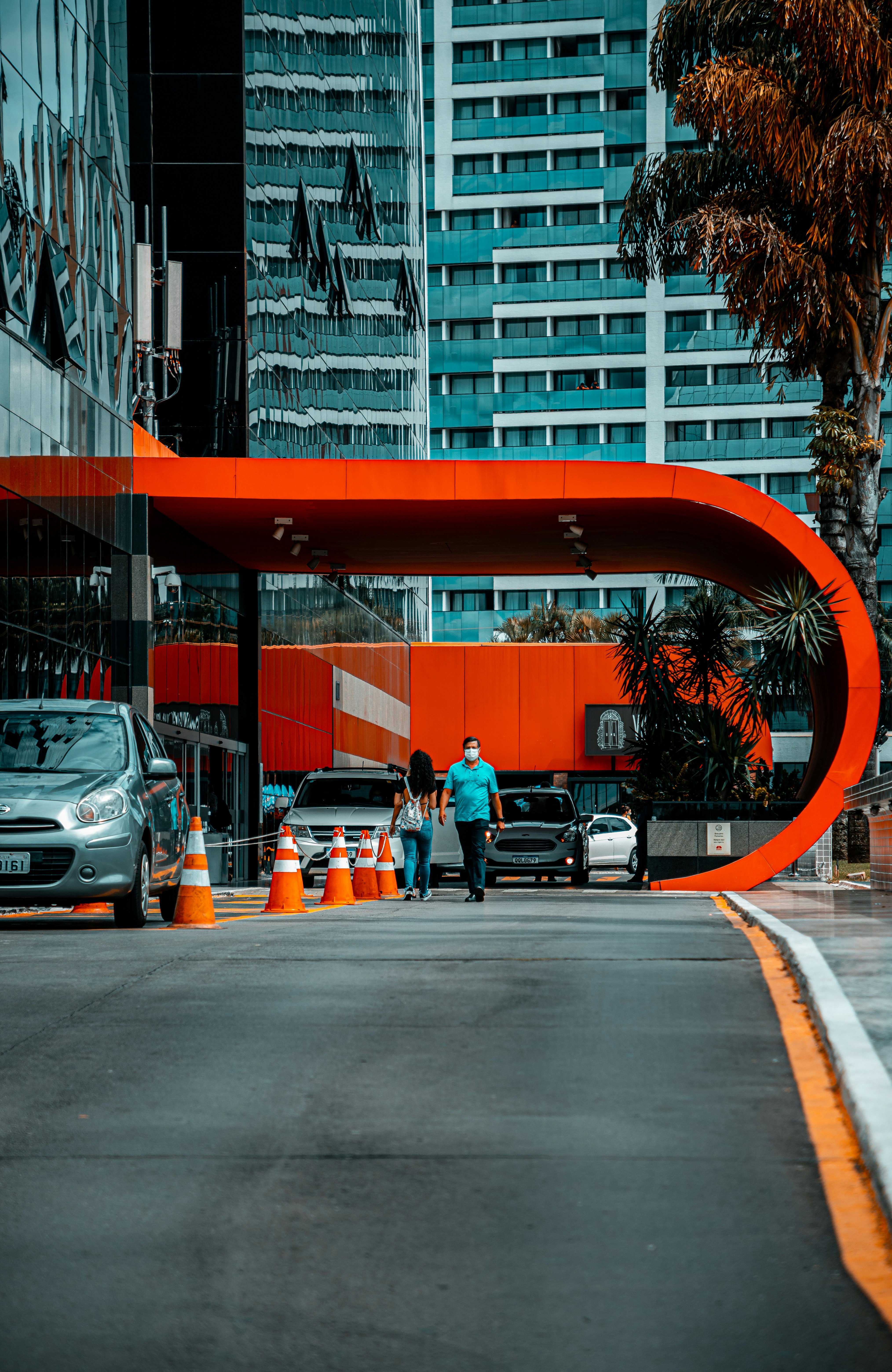 Two pedestrians navigate a vibrant urban pathway, framed by modern architecture and bold orange accents. Traffic cones add a playful touch to the scene.