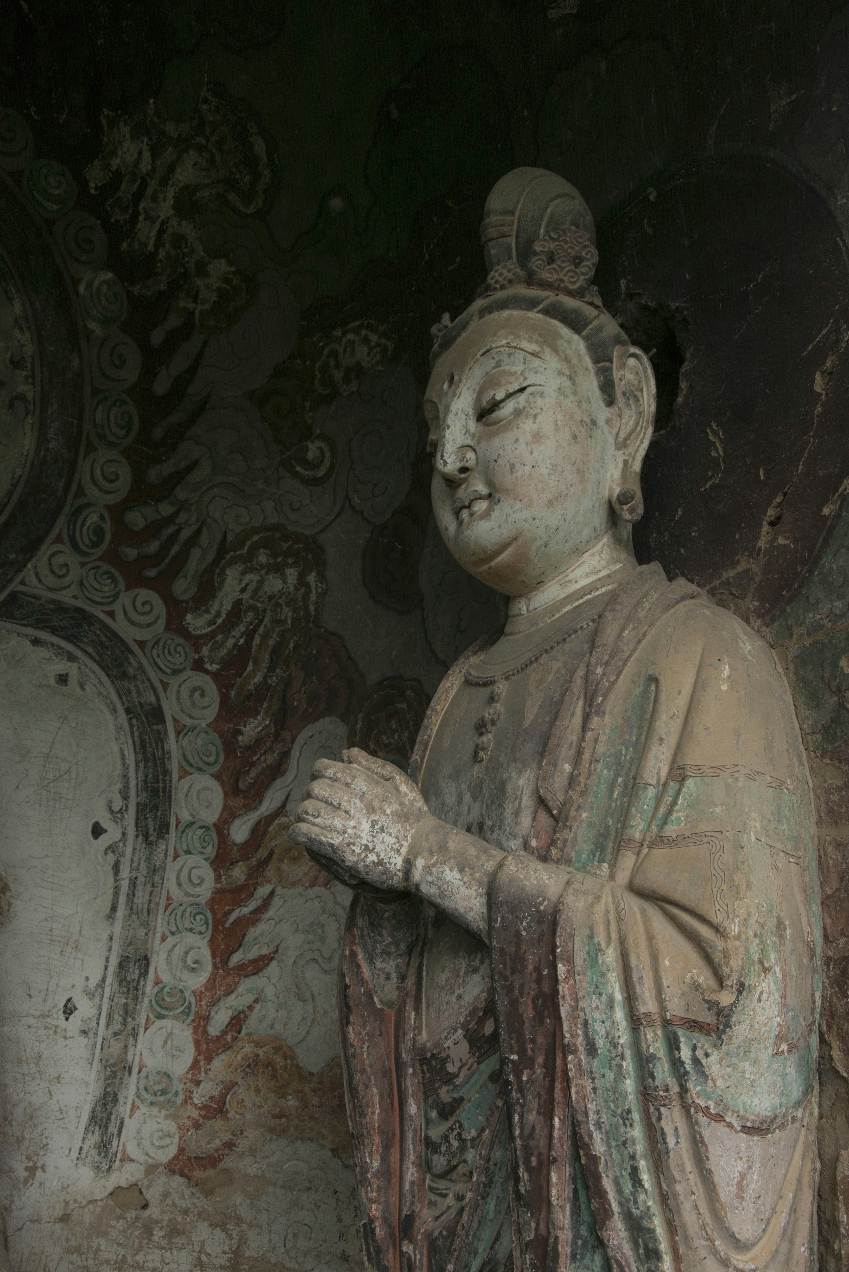 Weathered statue of a serene figure with clasped hands, set against a richly textured mural background.