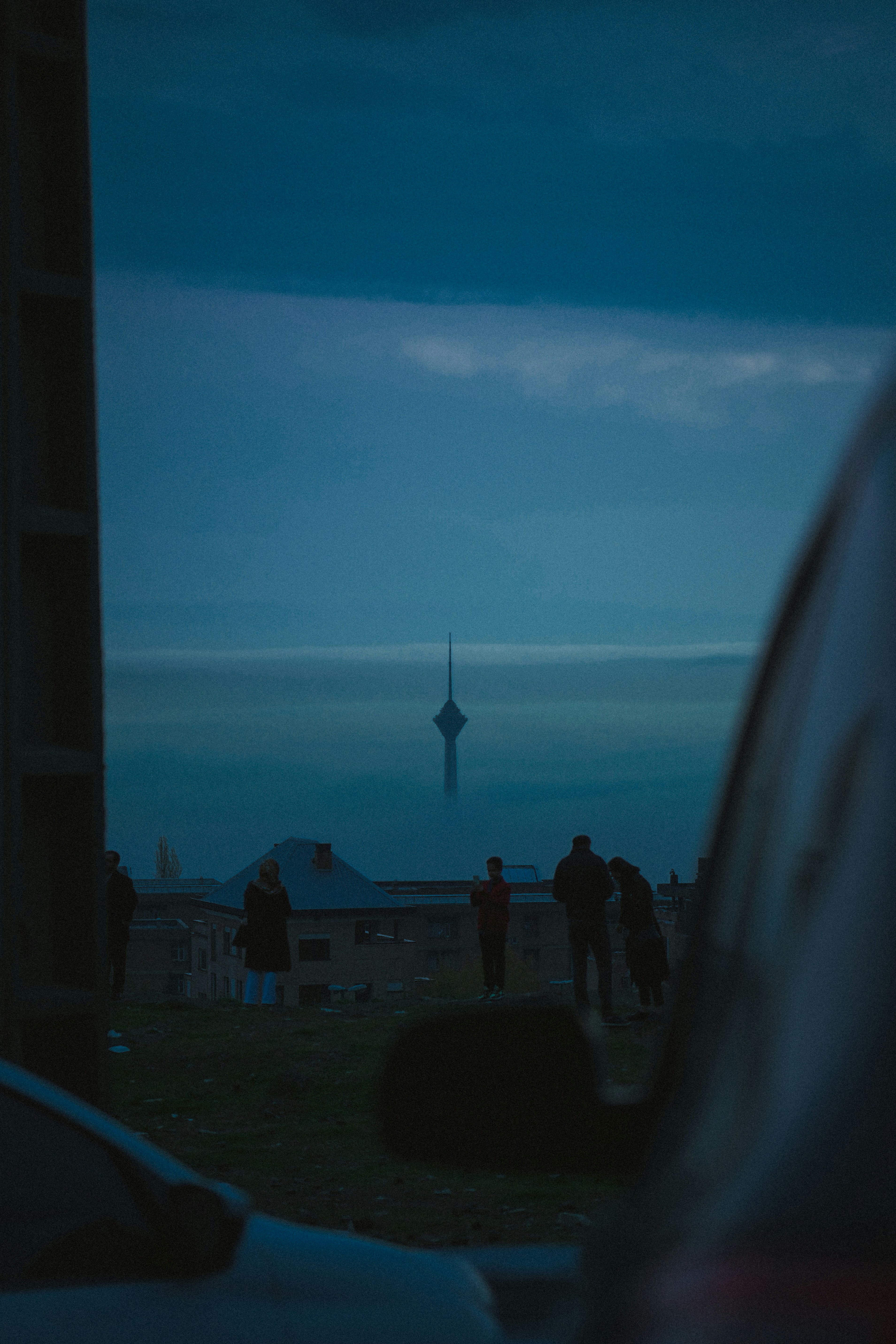 A towering structure looms in the distance, shrouded in mist, while silhouettes of people gather in the foreground. The scene evokes a sense of mystery and quiet contemplation.