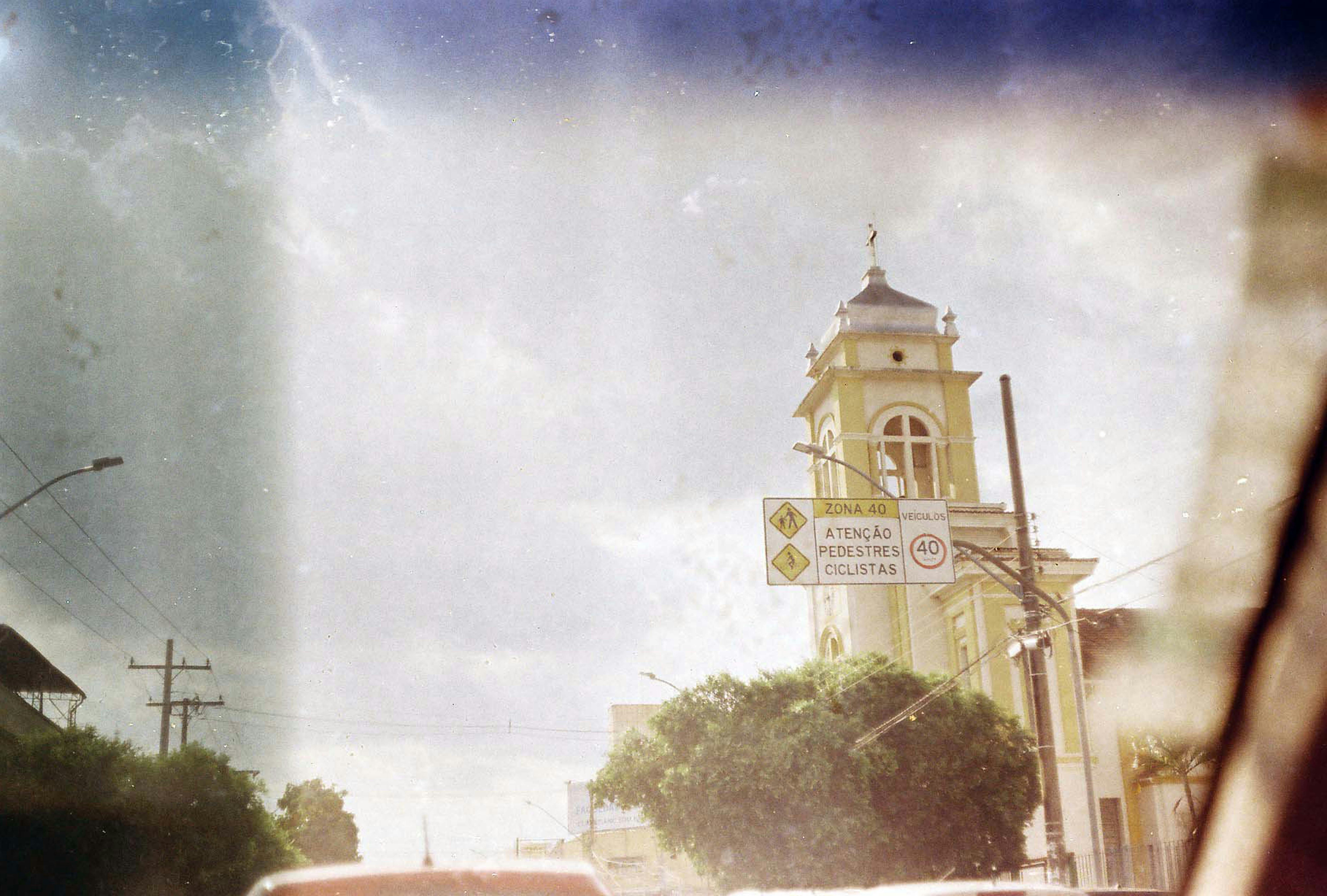 Yellow clock tower with a dome top stands against a partly cloudy sky, framed by blurred vehicles and urban elements.