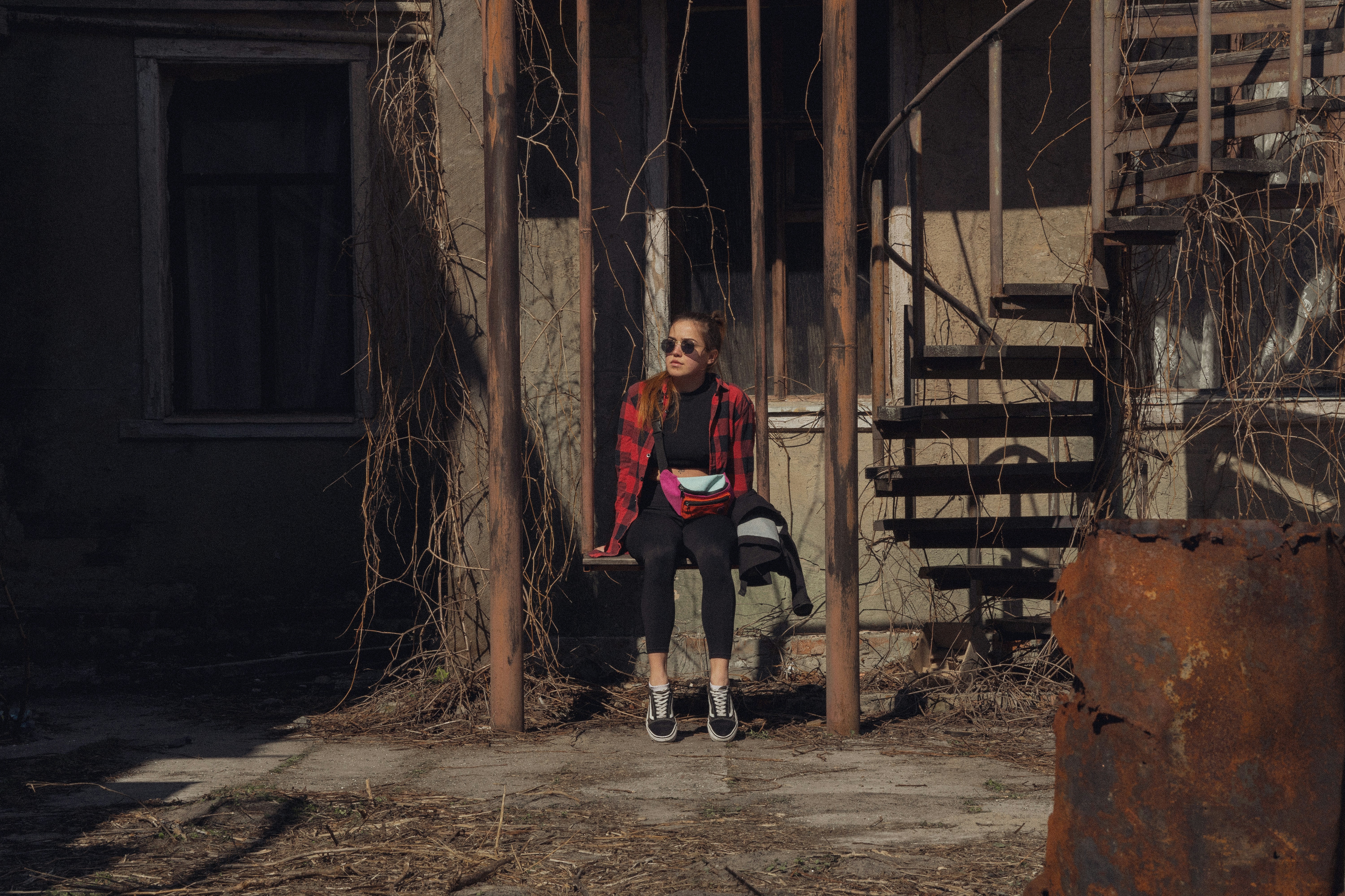 A young woman sits contemplatively on a spiral staircase in a derelict building, surrounded by creeping vines and remnants of rusted metal. The scene evokes a sense of nostalgia and isolation.