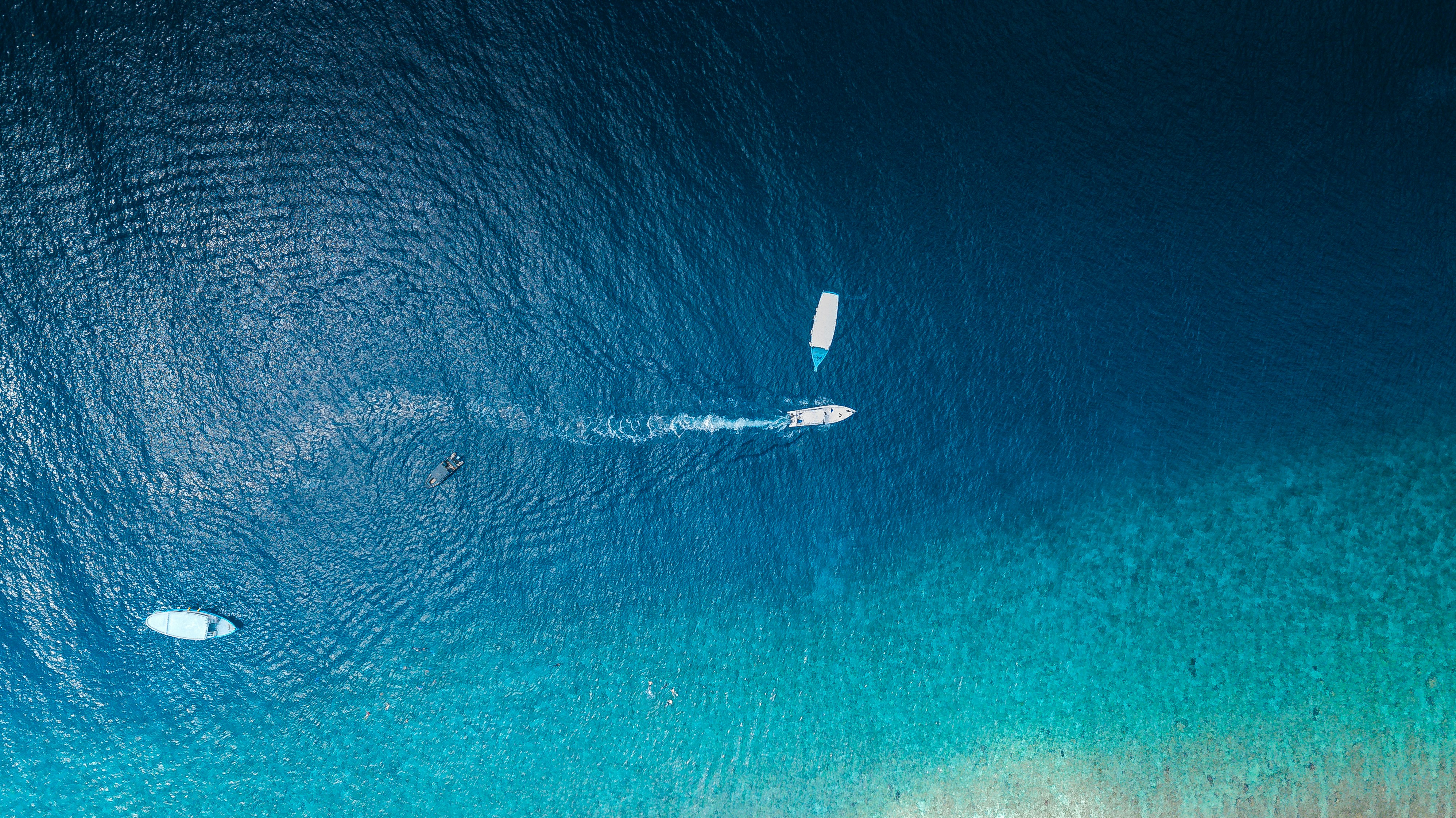 Aerial view of three boats creating swirling patterns in vibrant blue sea.