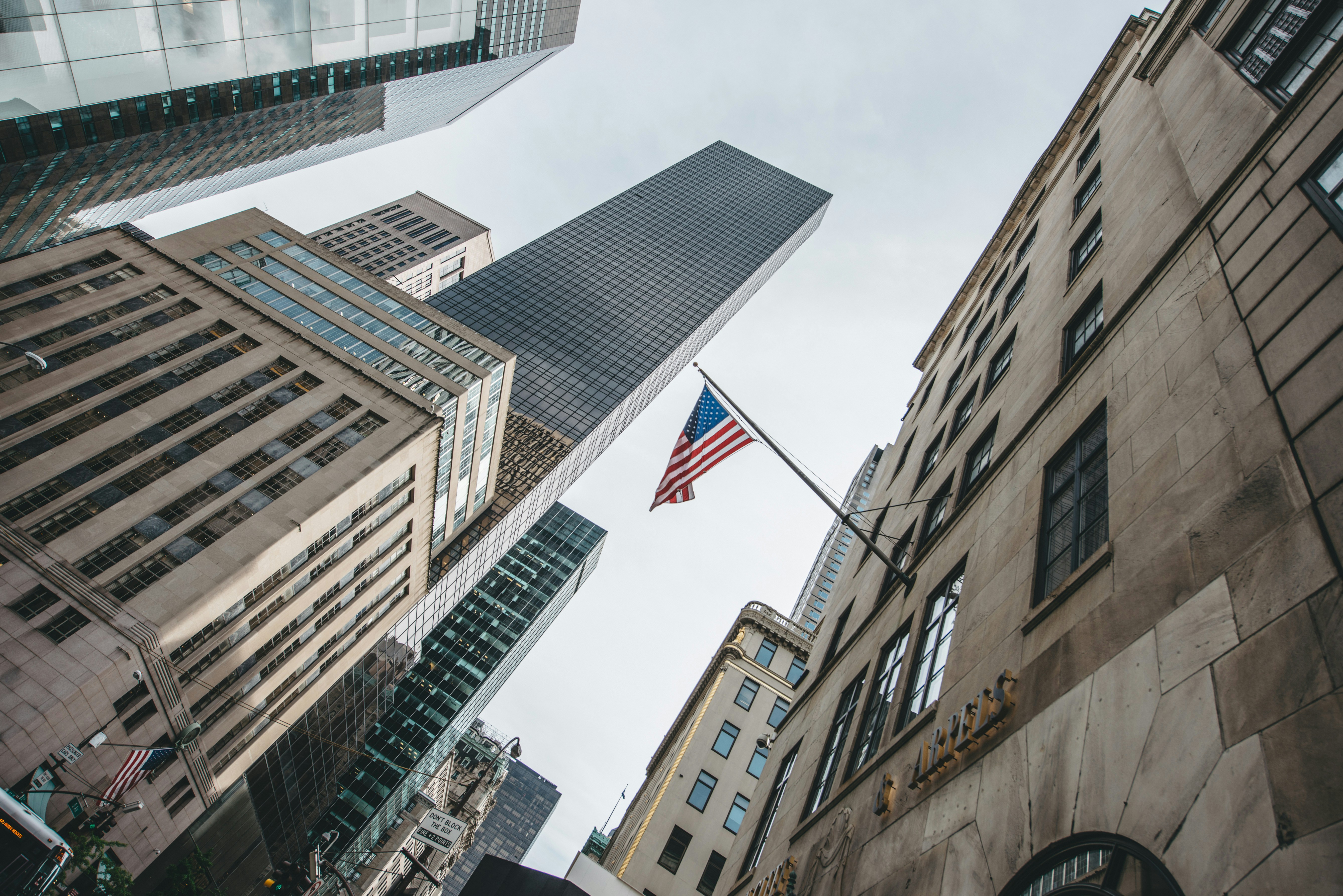 Skyscrapers converge in a dynamic composition, with an American flag prominently displayed against a cloudy backdrop.